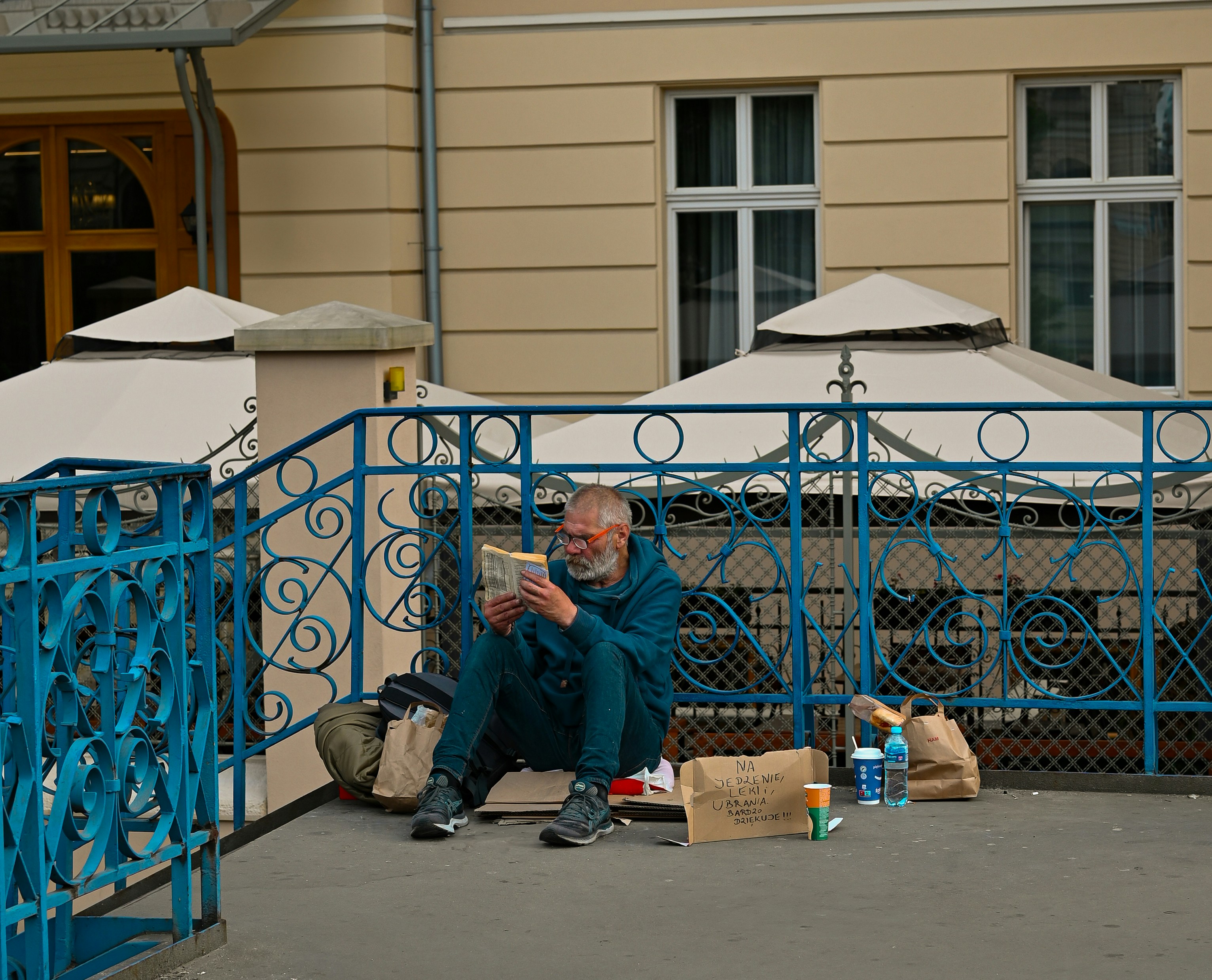 Homeless man reading a book on a city street in Kraków, sitting by a blue railing with a cardboard sign asking for help.