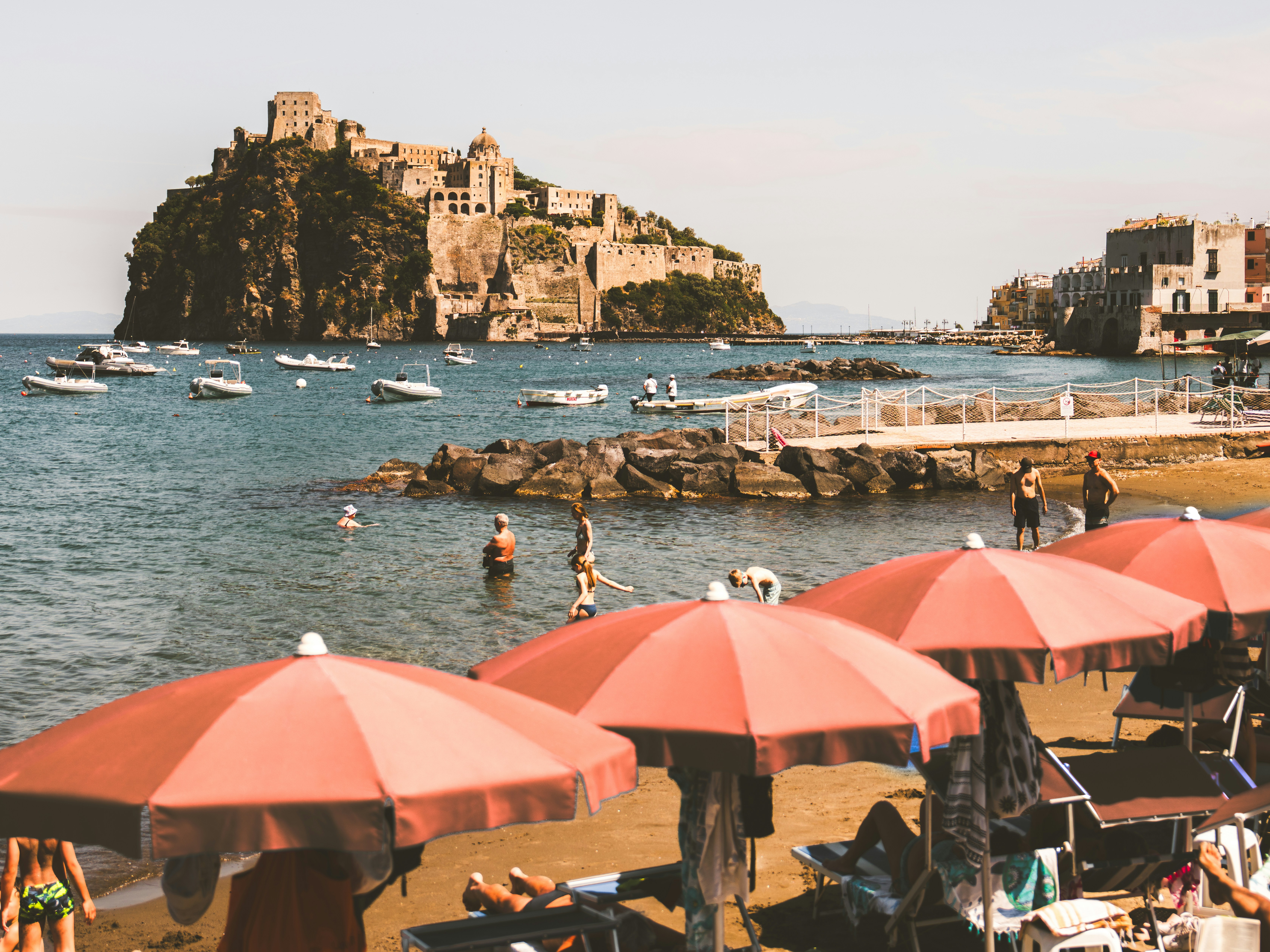 Beach with red umbrellas, swimmers, boats, and a castle island.