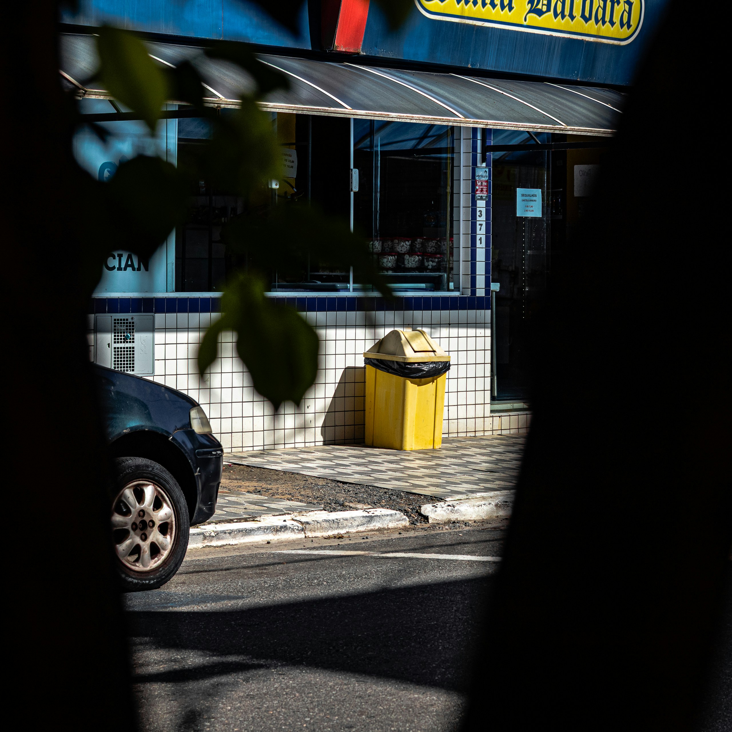 A lone yellow trash can sits on a tiled sidewalk in front of a store.