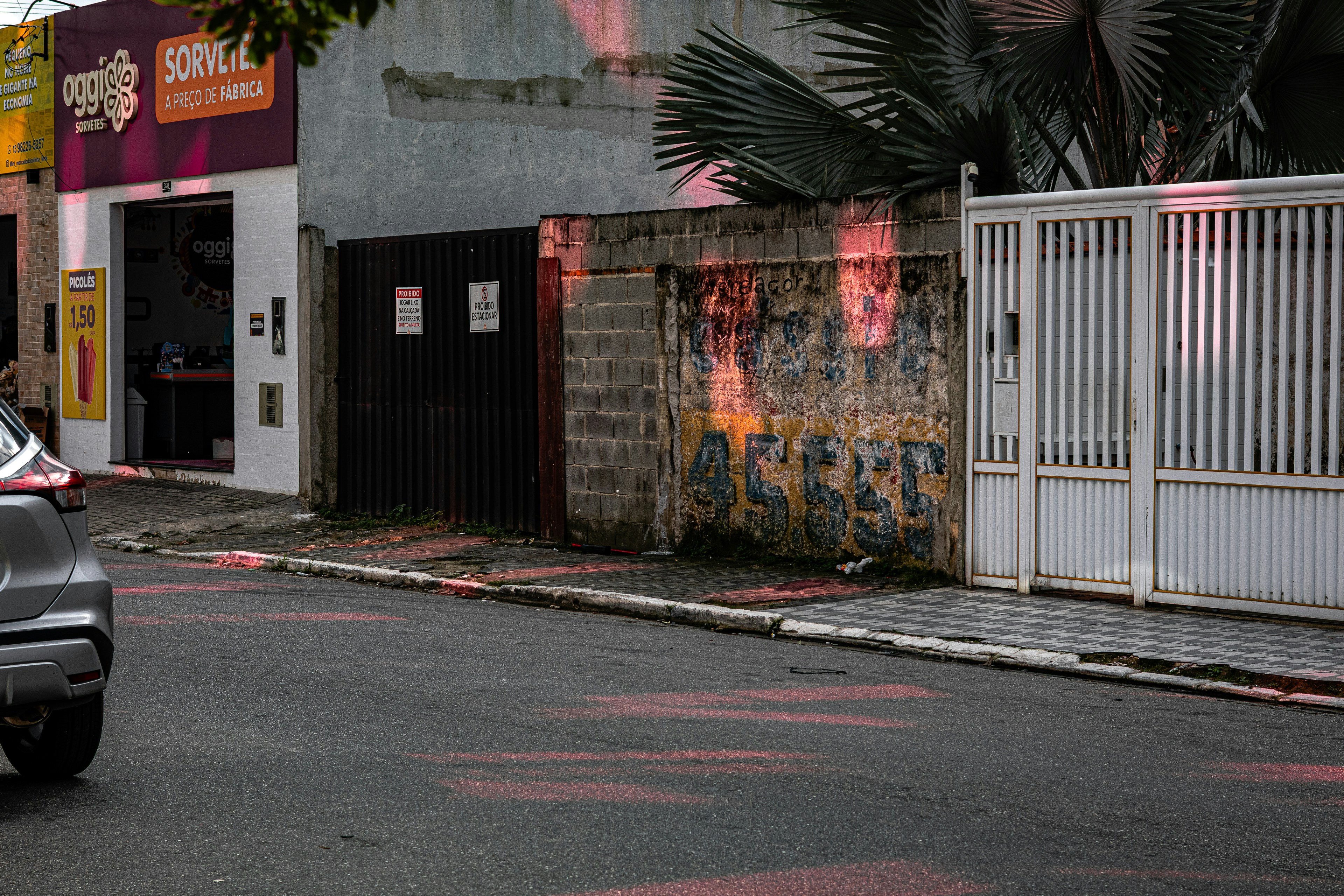 Graffiti and red light reflections on a street in front of a sorveteria and a white gate.