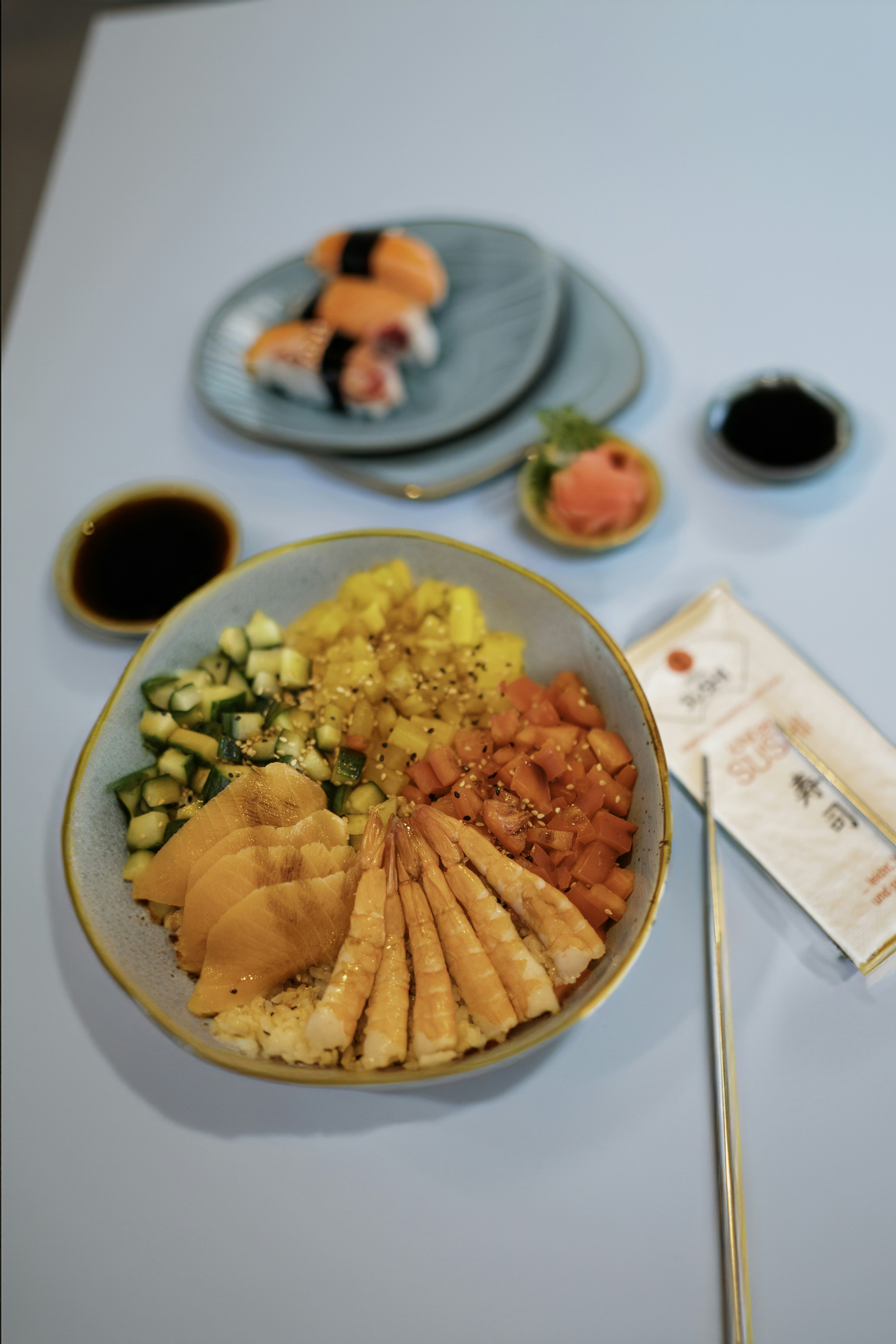 A colorful poke bowl with sushi, soy sauce, and chopsticks.