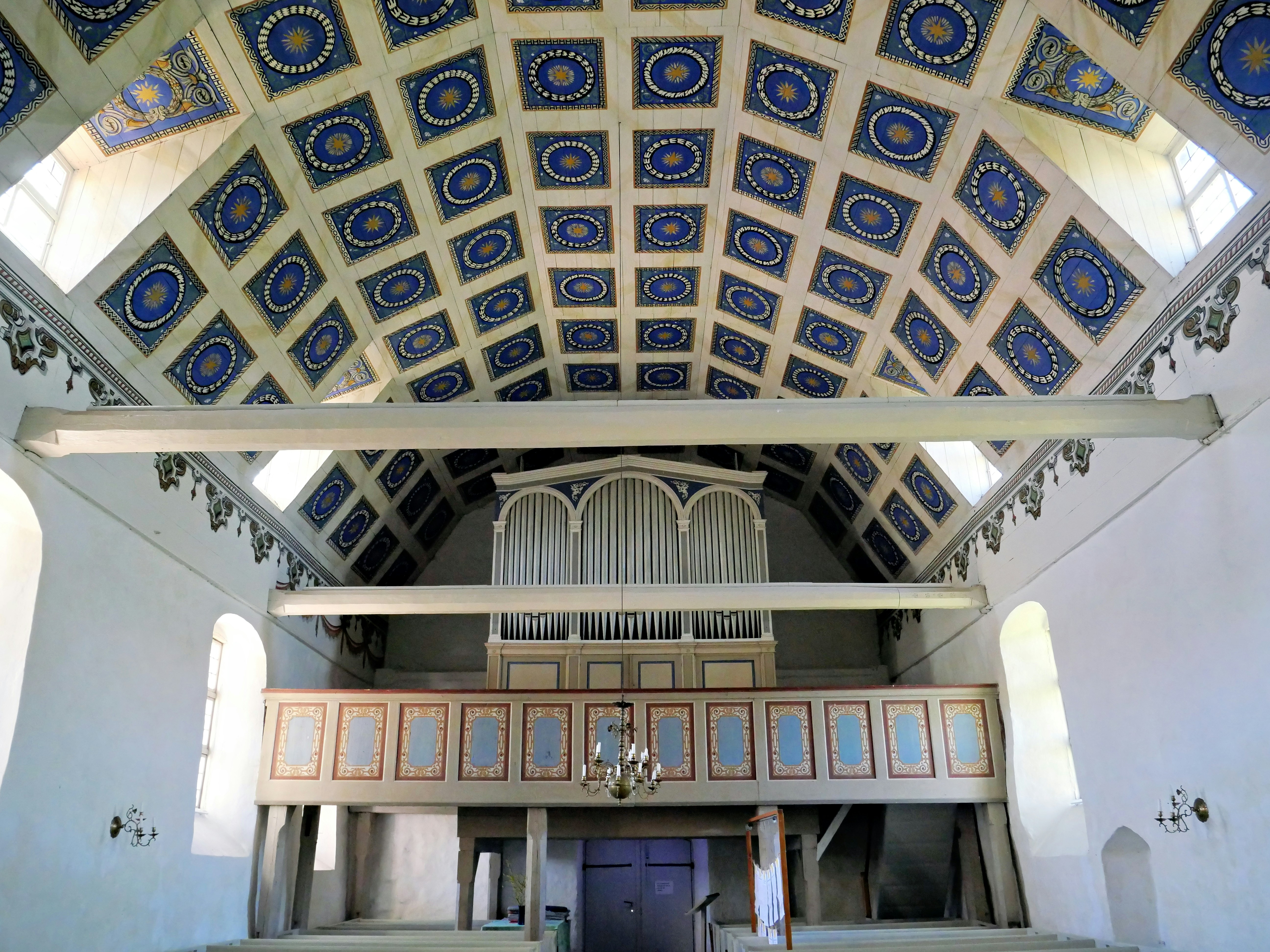 Historic interior featuring a decorative coffered ceiling and a high organ loft with paneling, plaster walls, and soft natural light from arched windows.