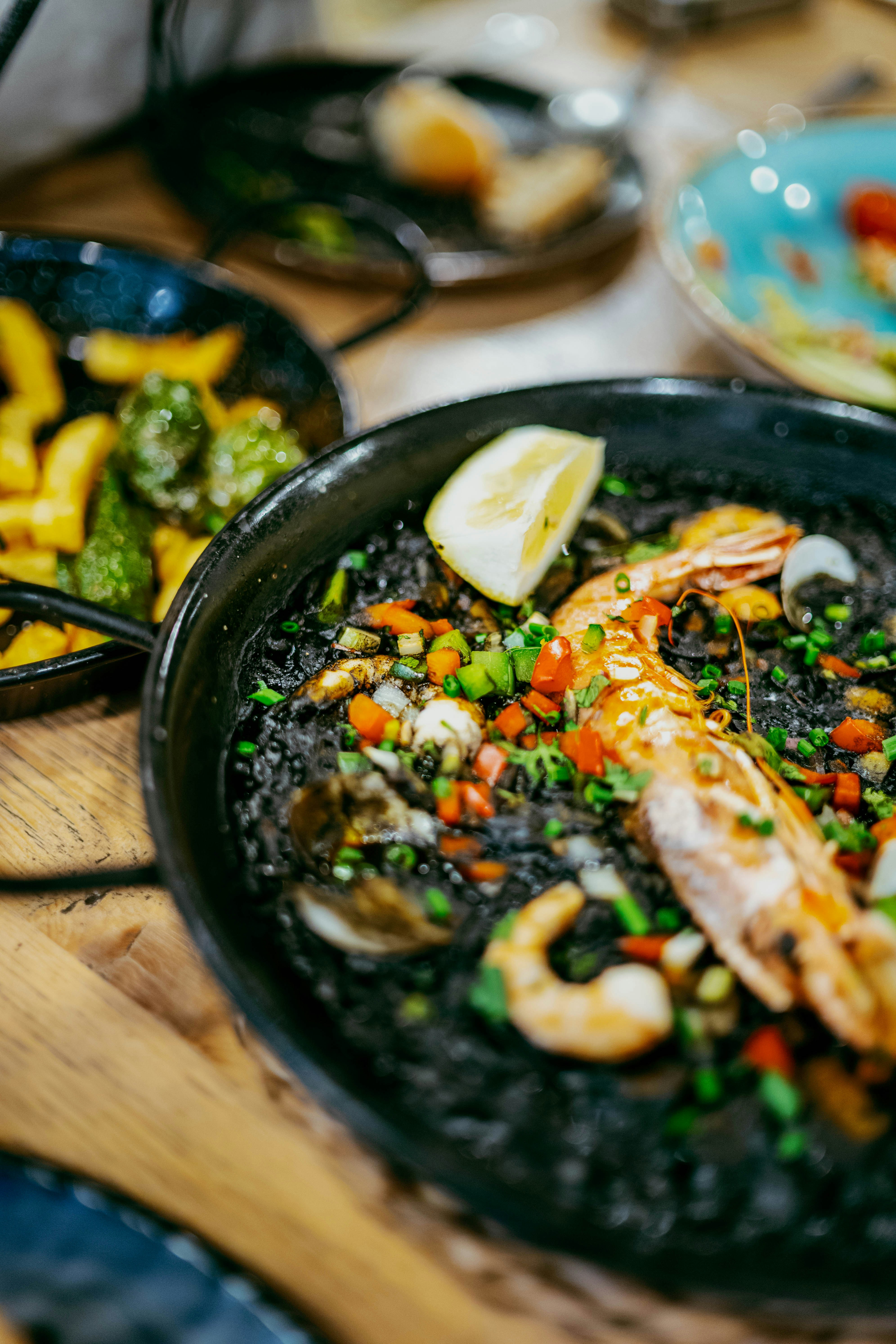 Black paella with shrimp, vegetables, and lemon on a table.