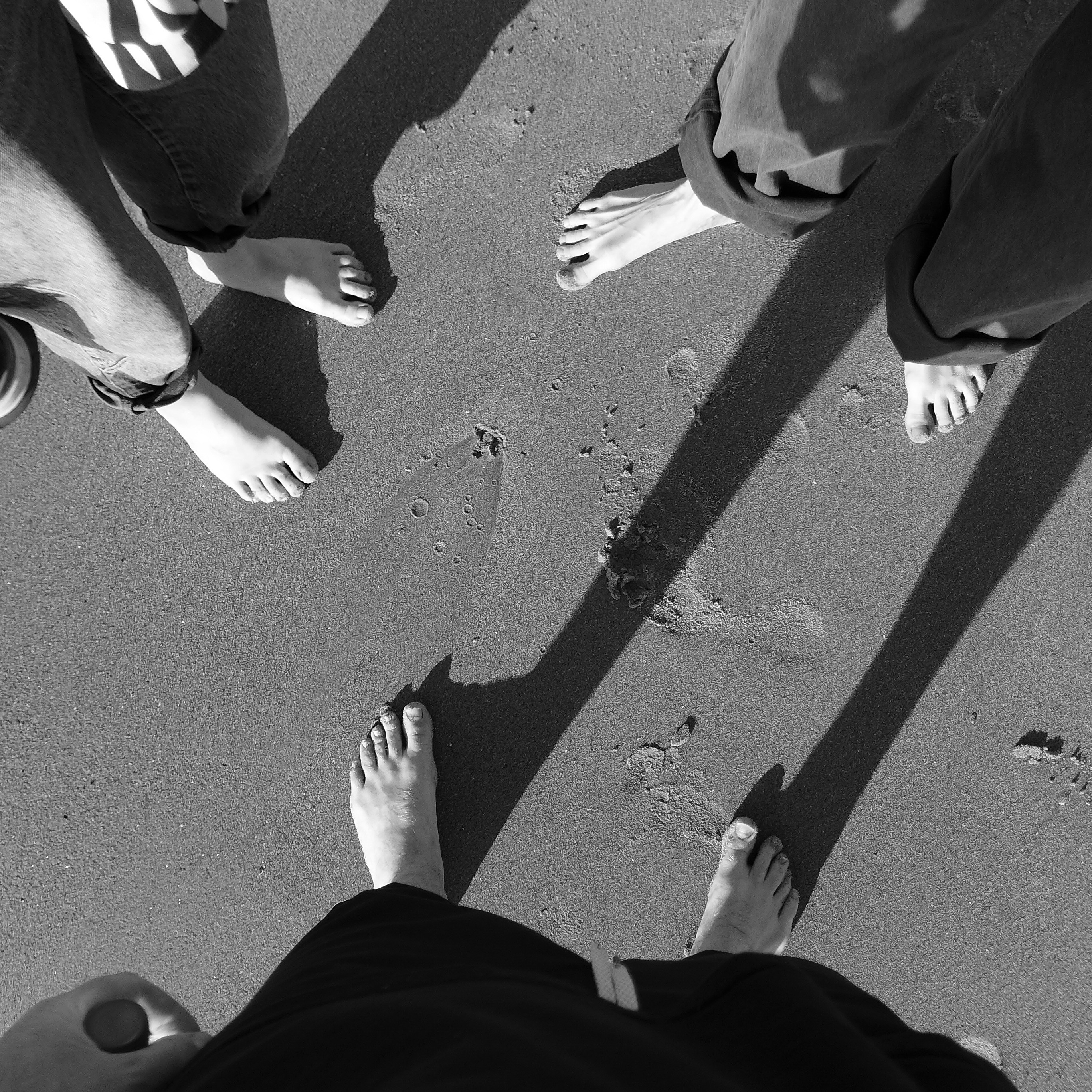 Bare feet of five people on a sandy beach, with long shadows and footprints creating radial composition. A candid, overhead view emphasizing interaction with the ground and a casual moment by the water's edge.