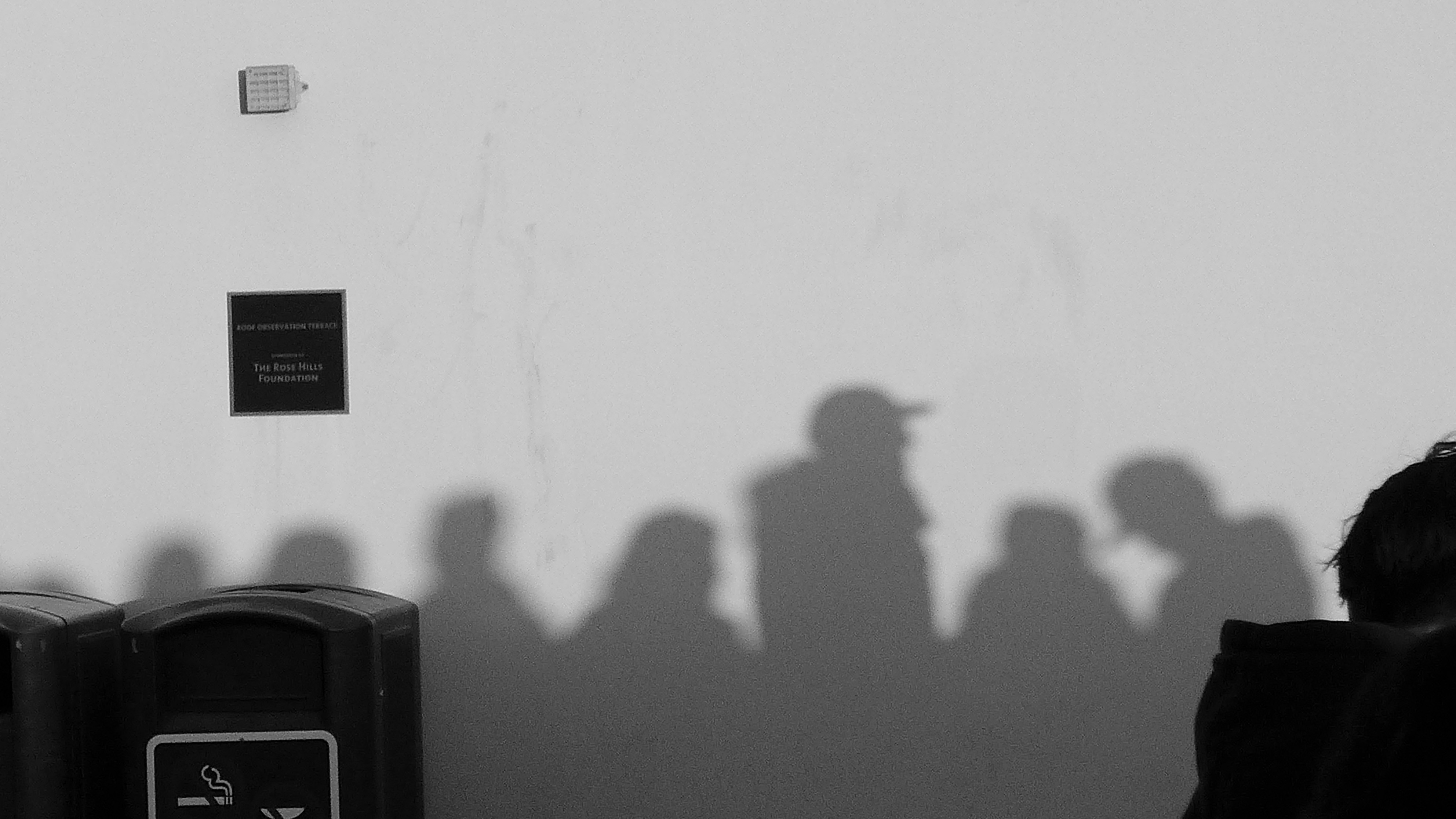 Monochrome scene of a wall with dark silhouettes of people projected or cast as shadows, with trash bins in the lower left. Subtle wall textures and signage visible, emphasizing urban ambience.