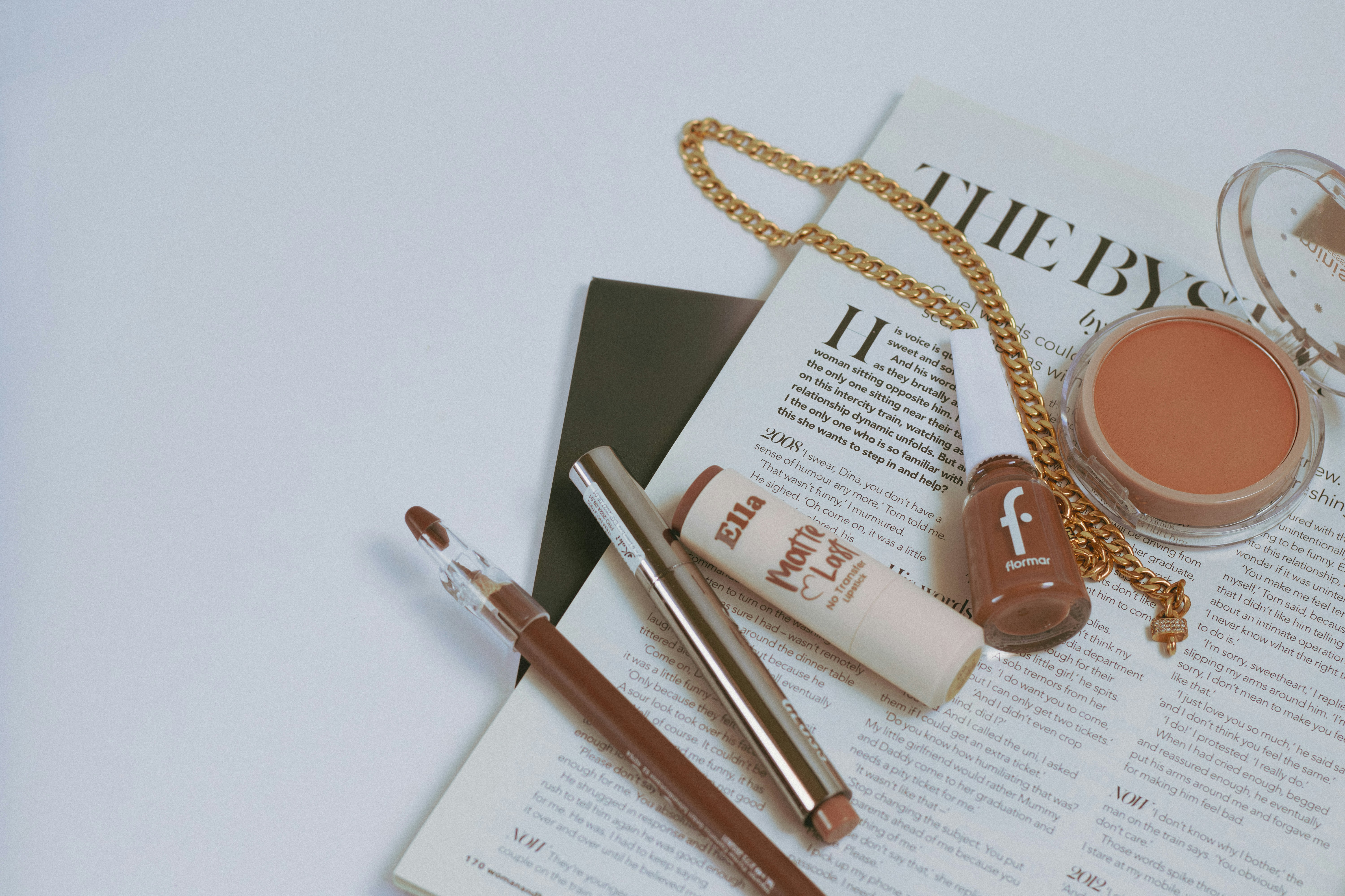 Various makeup products, magazine, and gold chain on white background.
