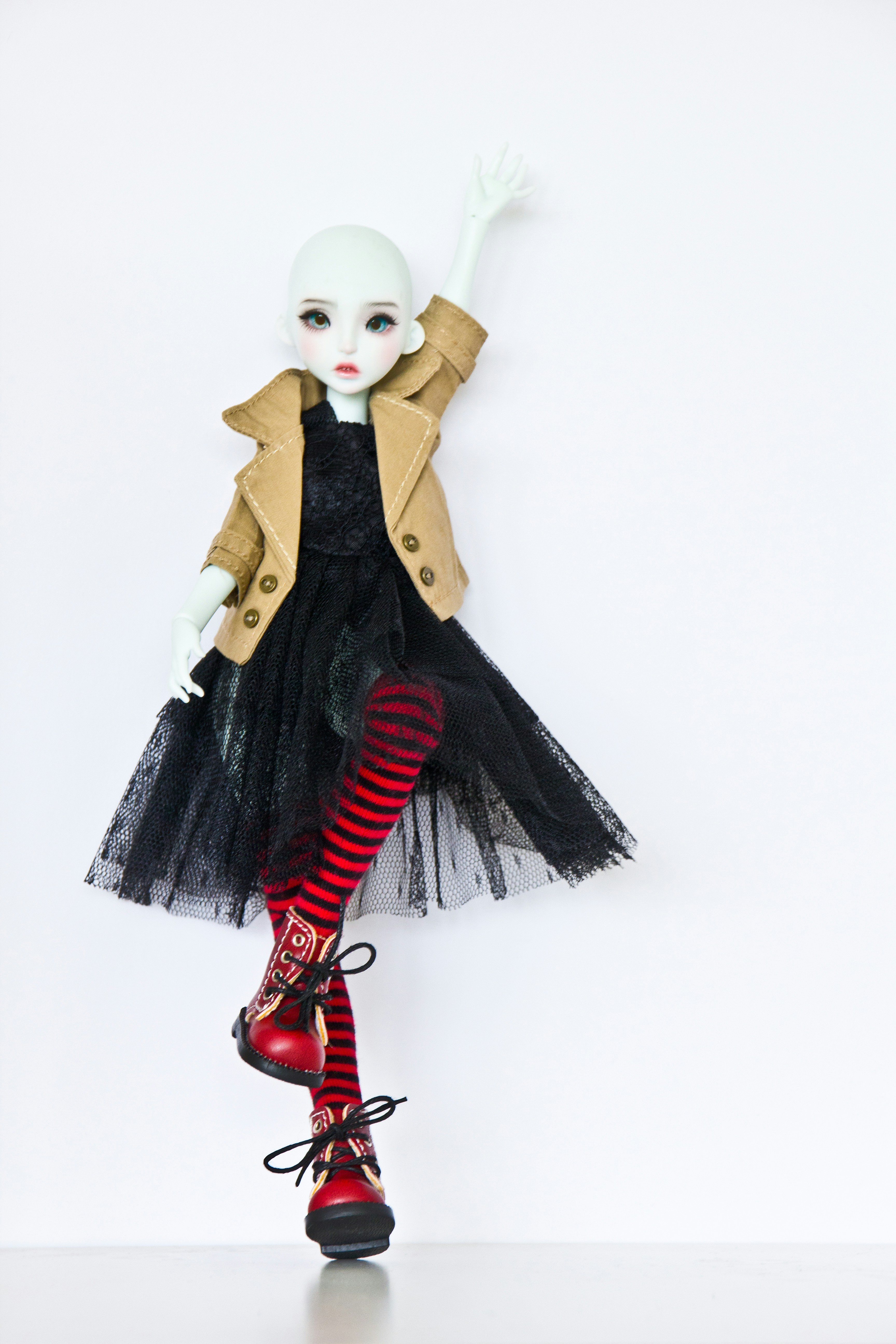 Doll in a Tan Trench and Red-Striped Tights Leaping Against a White Backdrop
