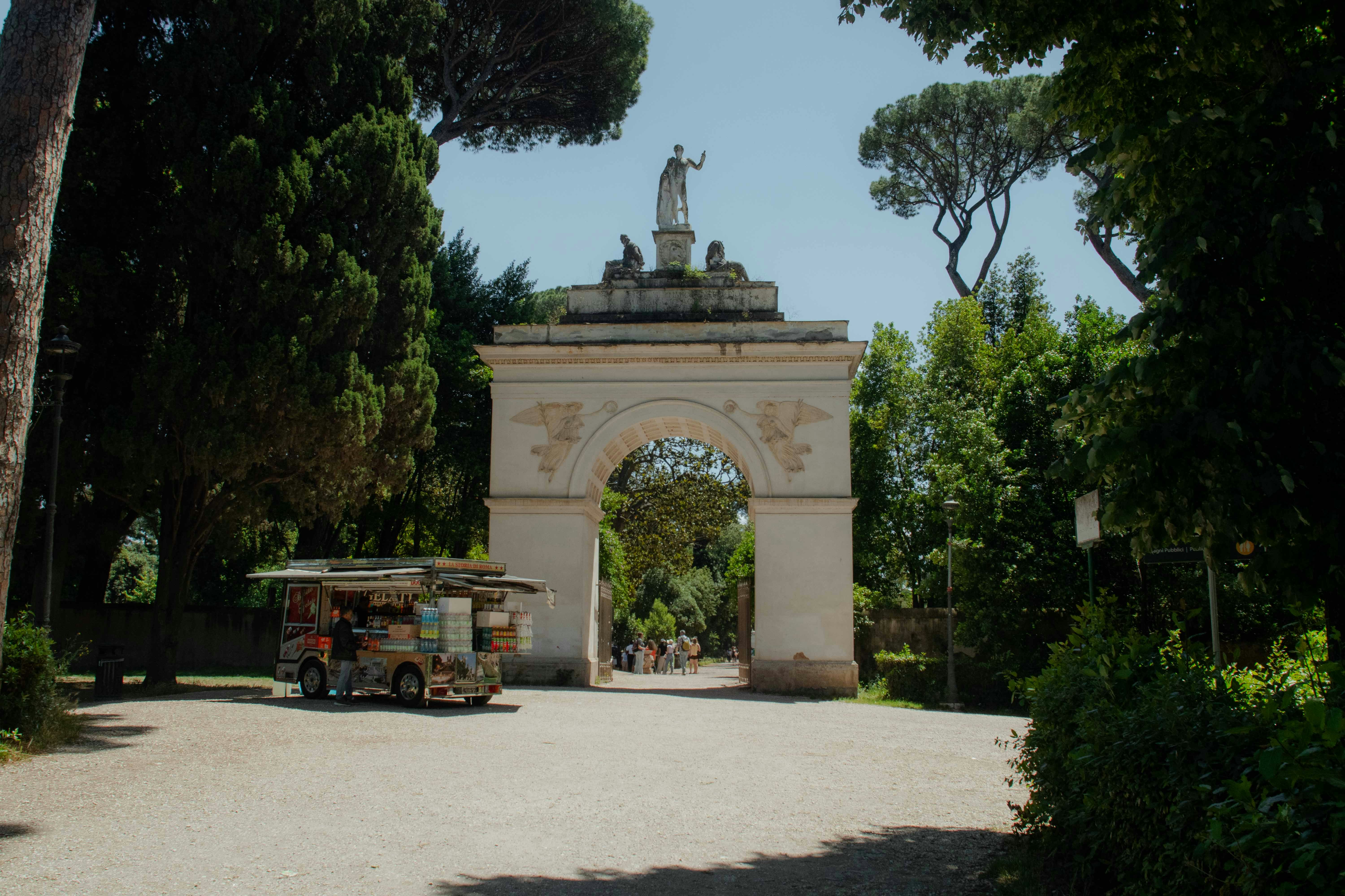 Archway of Vue: A sunlit park entrance guarded by trees and a small food cart