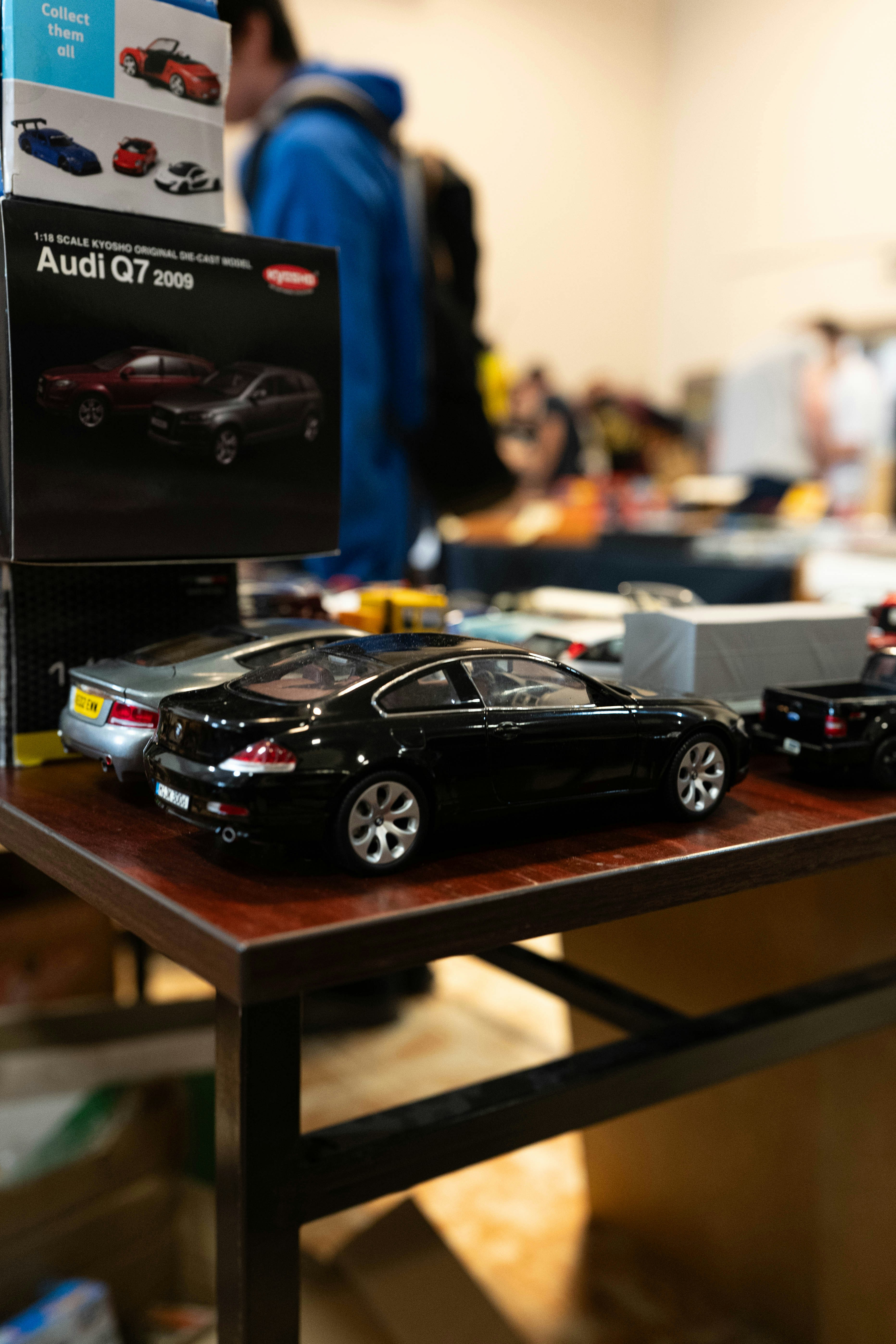 Miniature car models displayed on a wooden table at a bustling exhibition, with blurred visitors in the background.