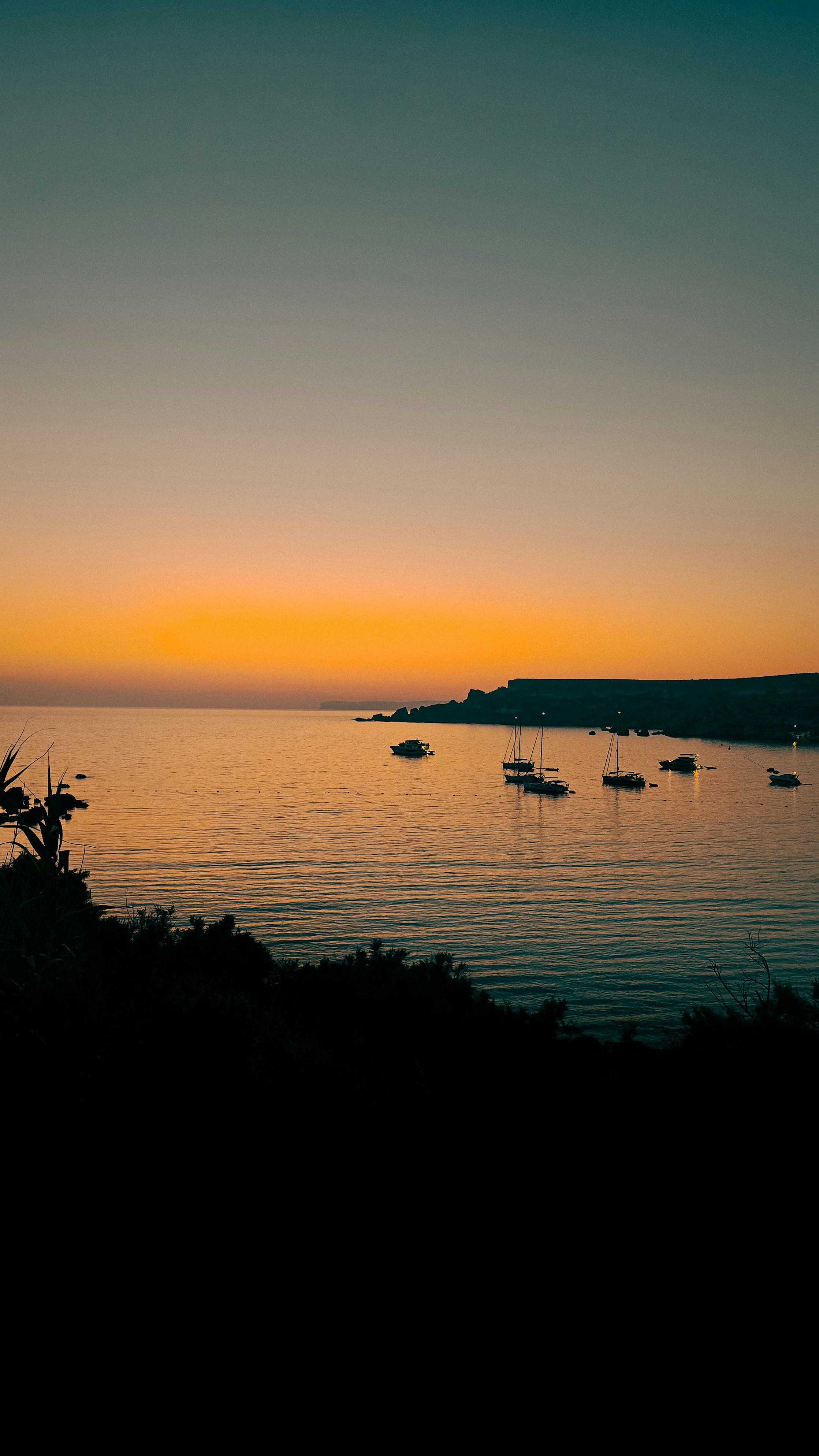 Sunset over a calm bay with boats and distant coastline.