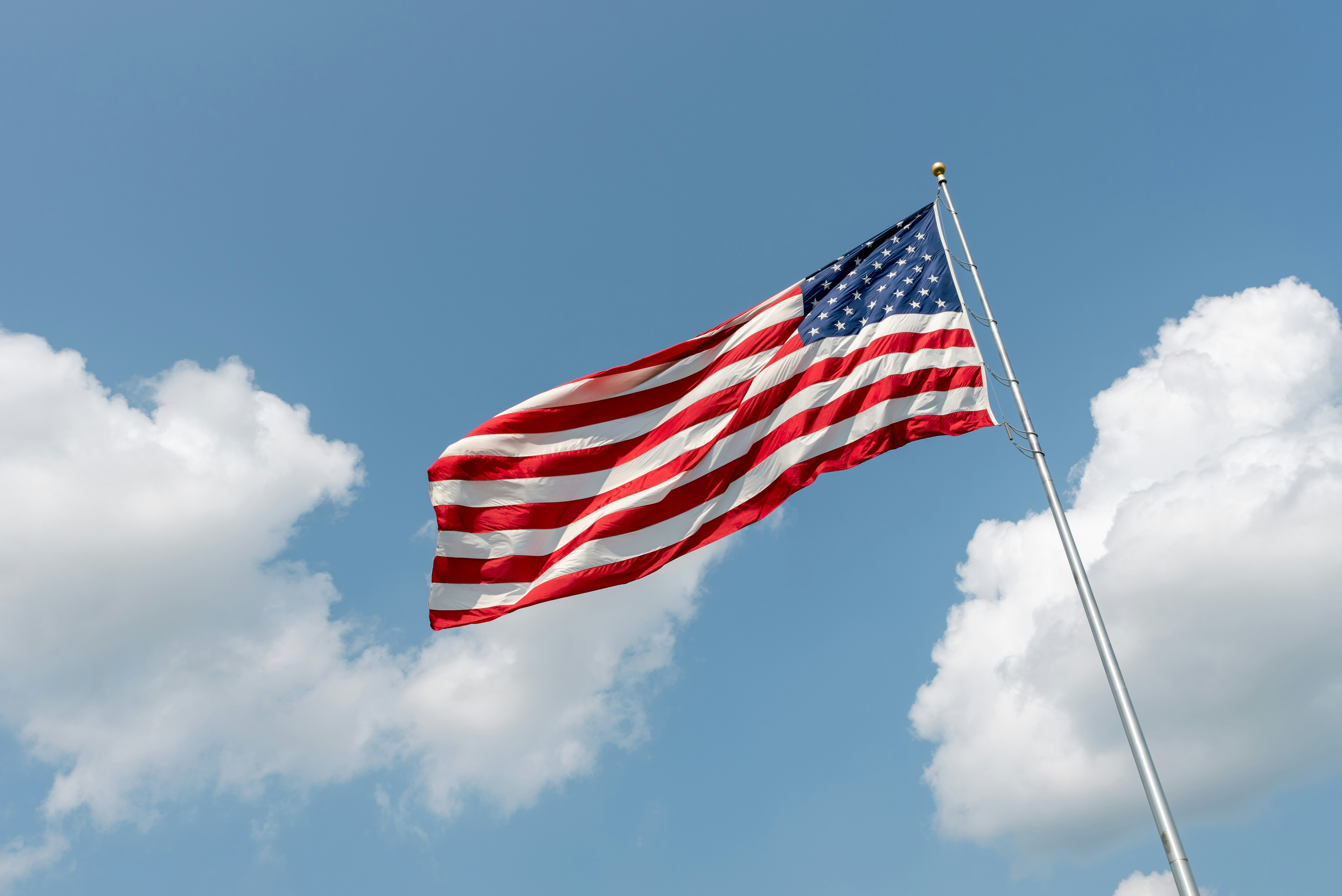 An American flag waves steadily from a silver flagpole in a clean, classically composed low-angle shot, with large billowing white cumulus clouds flanking both sides of the flag against a vivid blue sky. The flag is fully extended and evenly lit, its red, white, and blue colors rendered with excellent clarity and saturation. The wider framing gives more breathing room around the flag, allowing the dramatic cloud formations to become an equal part of the composition and adding a sense of grandeur and open sky. Perfect for patriotism, national holidays, civic pride, government, military, Independence Day, or clean versatile Americana concepts.