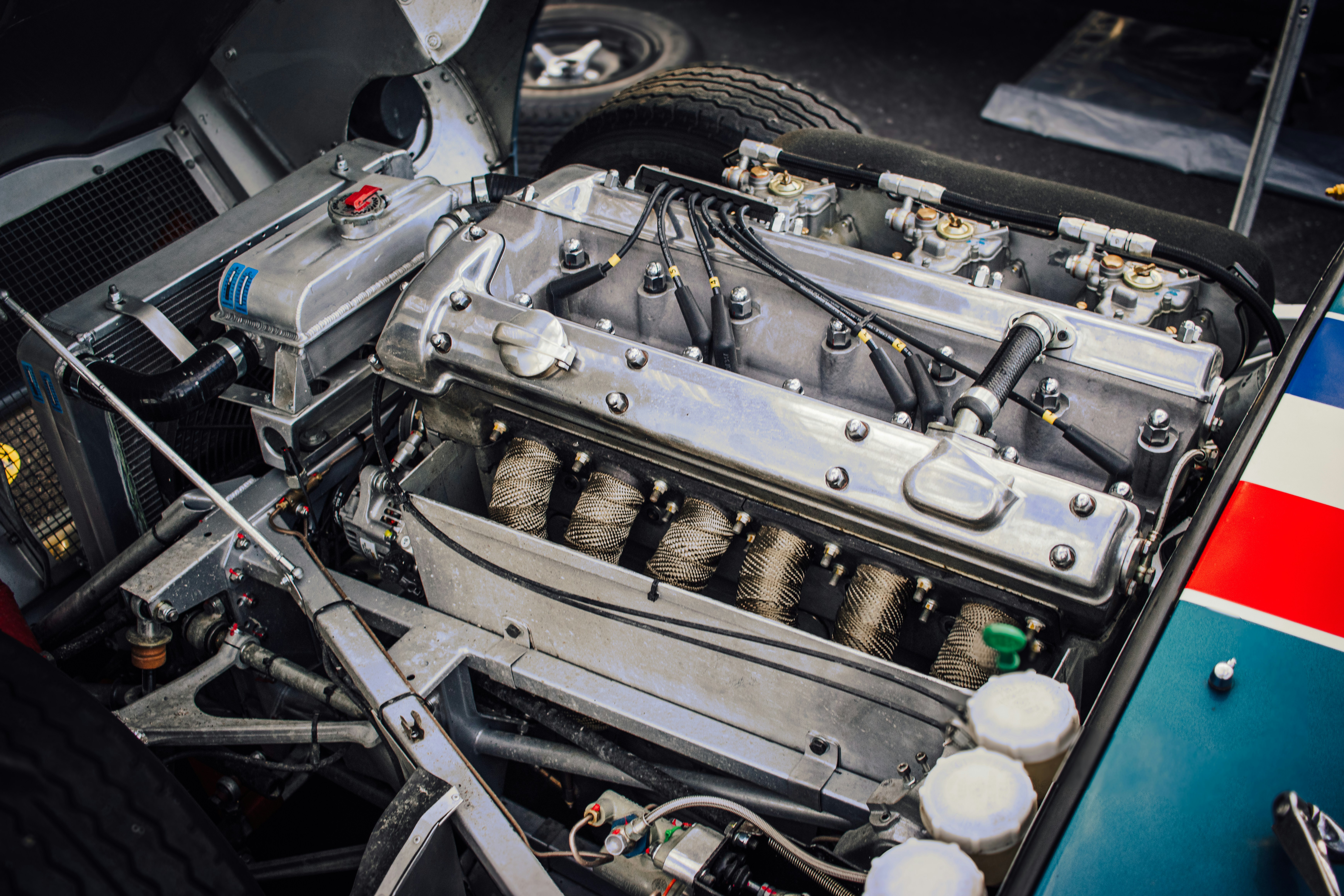 Close-up of a vintage inline-6 engine in a race car, chrome and aluminum components gleaming amid cockpit plumbing and battery loom.