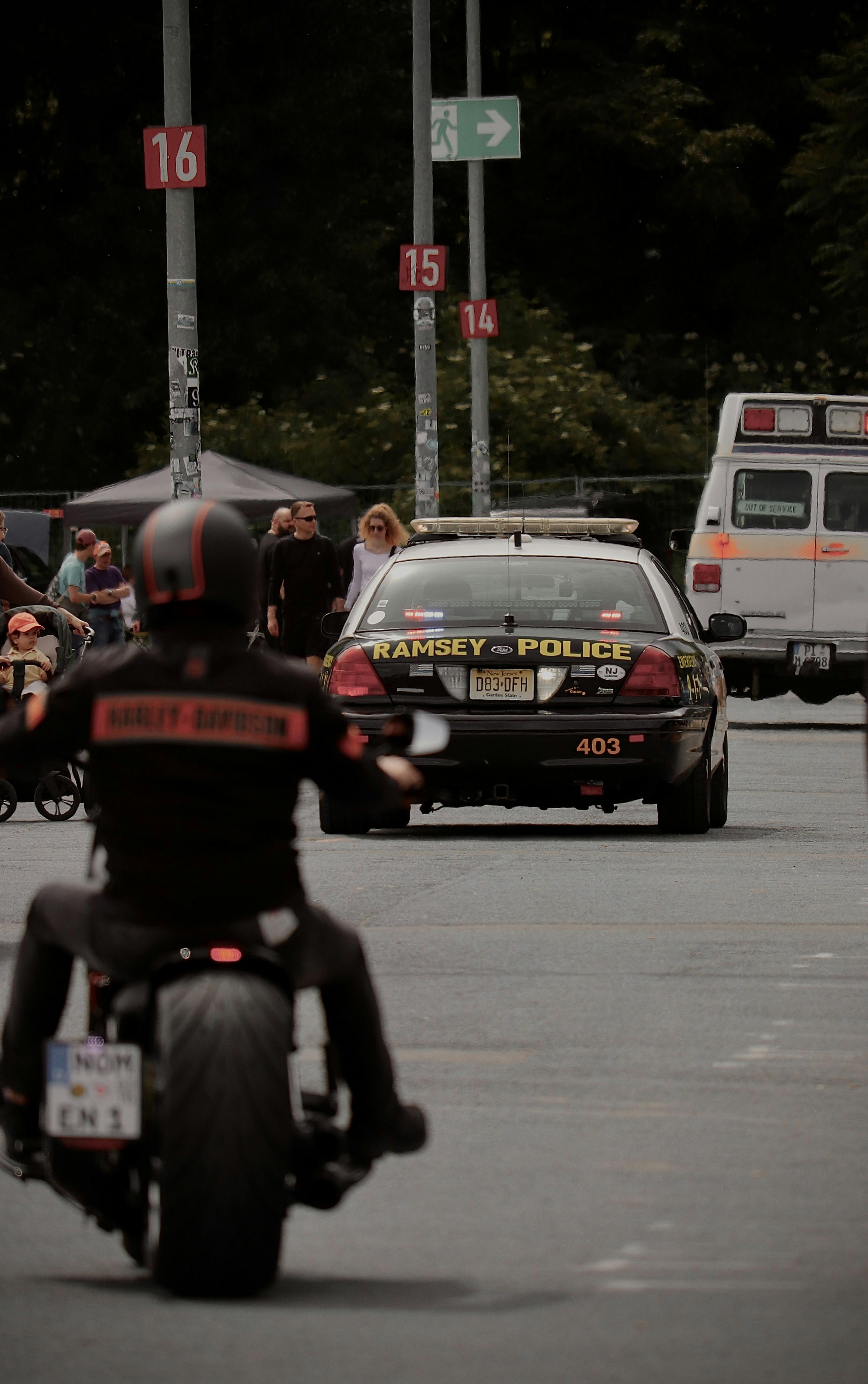 A Ramsey Police car advances toward the foreground after a street meetup, with a motorcyclist in the foreground and an ambulance on the right; the scene unfolds under numbered posts in an urban plaza.