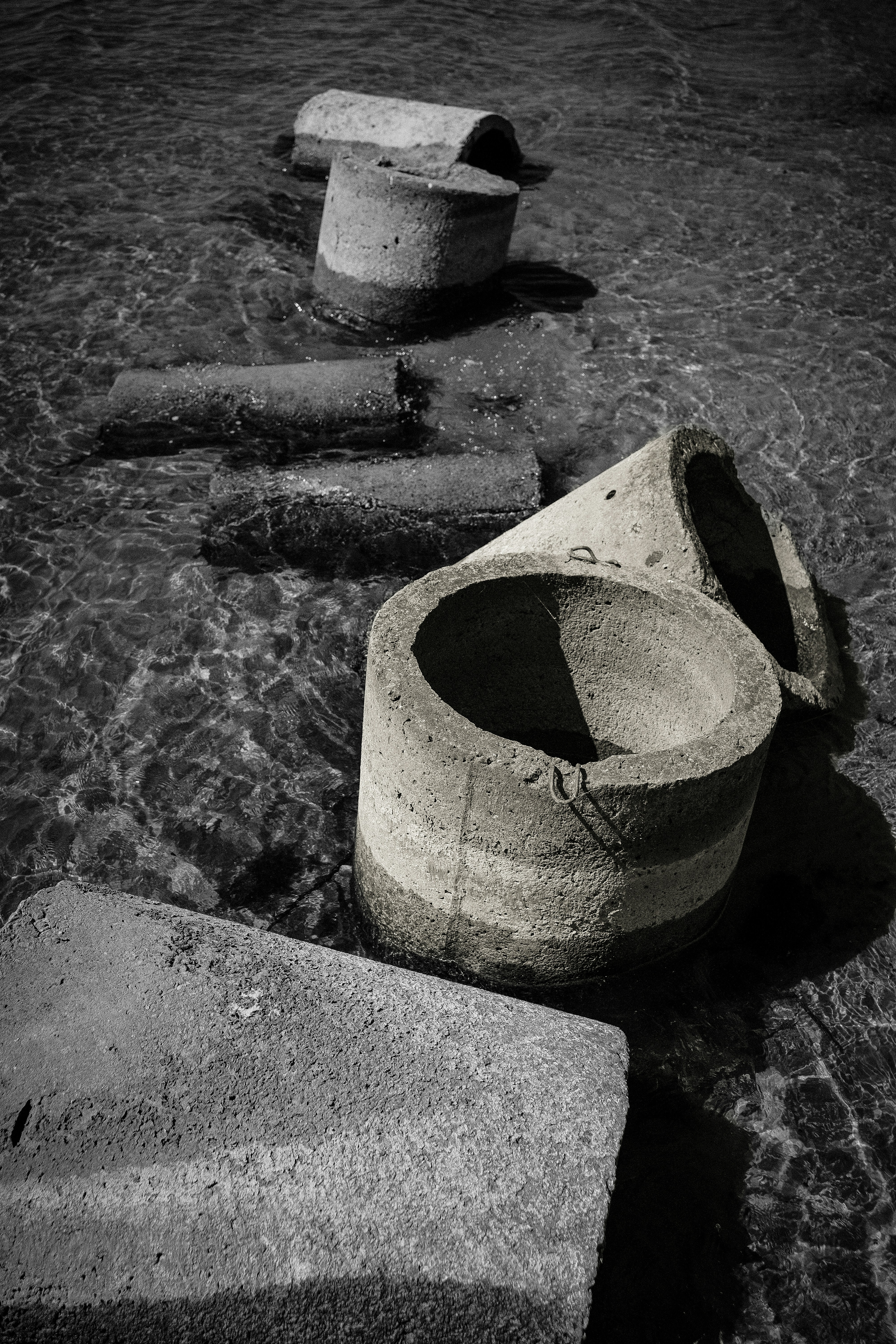 Monochrome shoreline scene with weathered concrete cylinders protruding from shallow water, forming a diagonal stack and casting textures on the rippled surface.