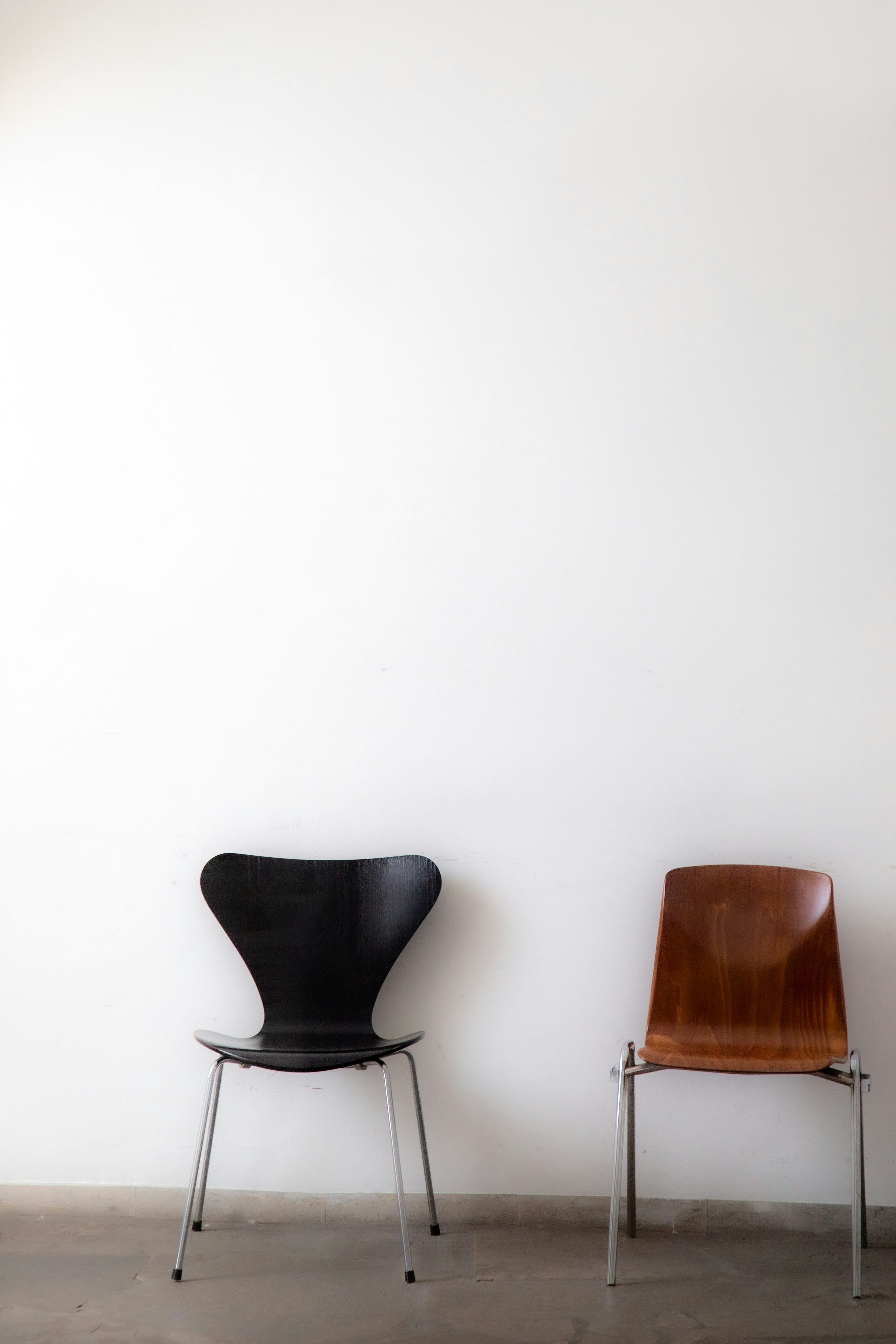 Two chairs sit against a stark white wall, one black and one wooden brown, creating a quiet, minimal interior scene.
