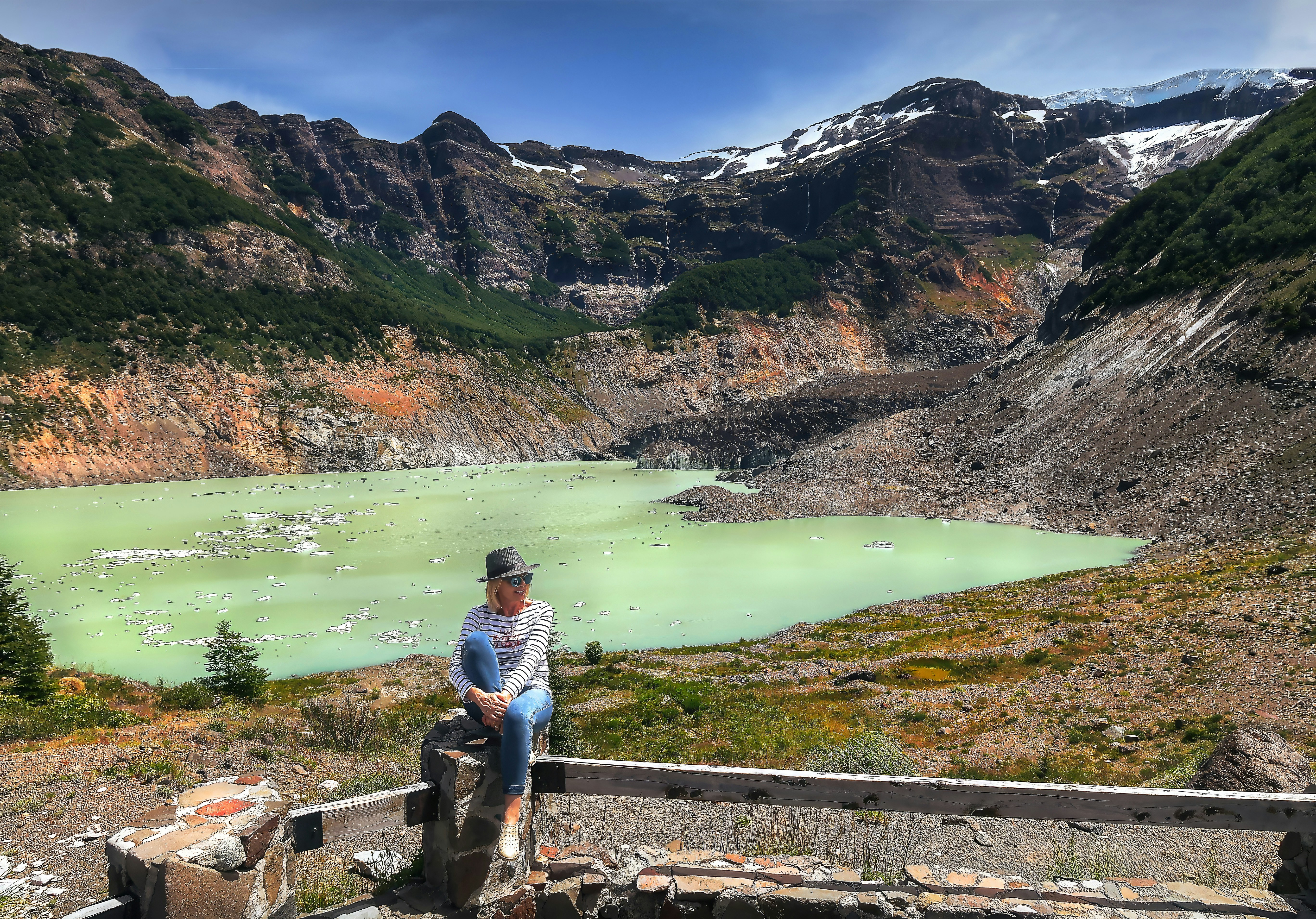 Woman sits by a vibrant glacial lake with snow-capped mountains.