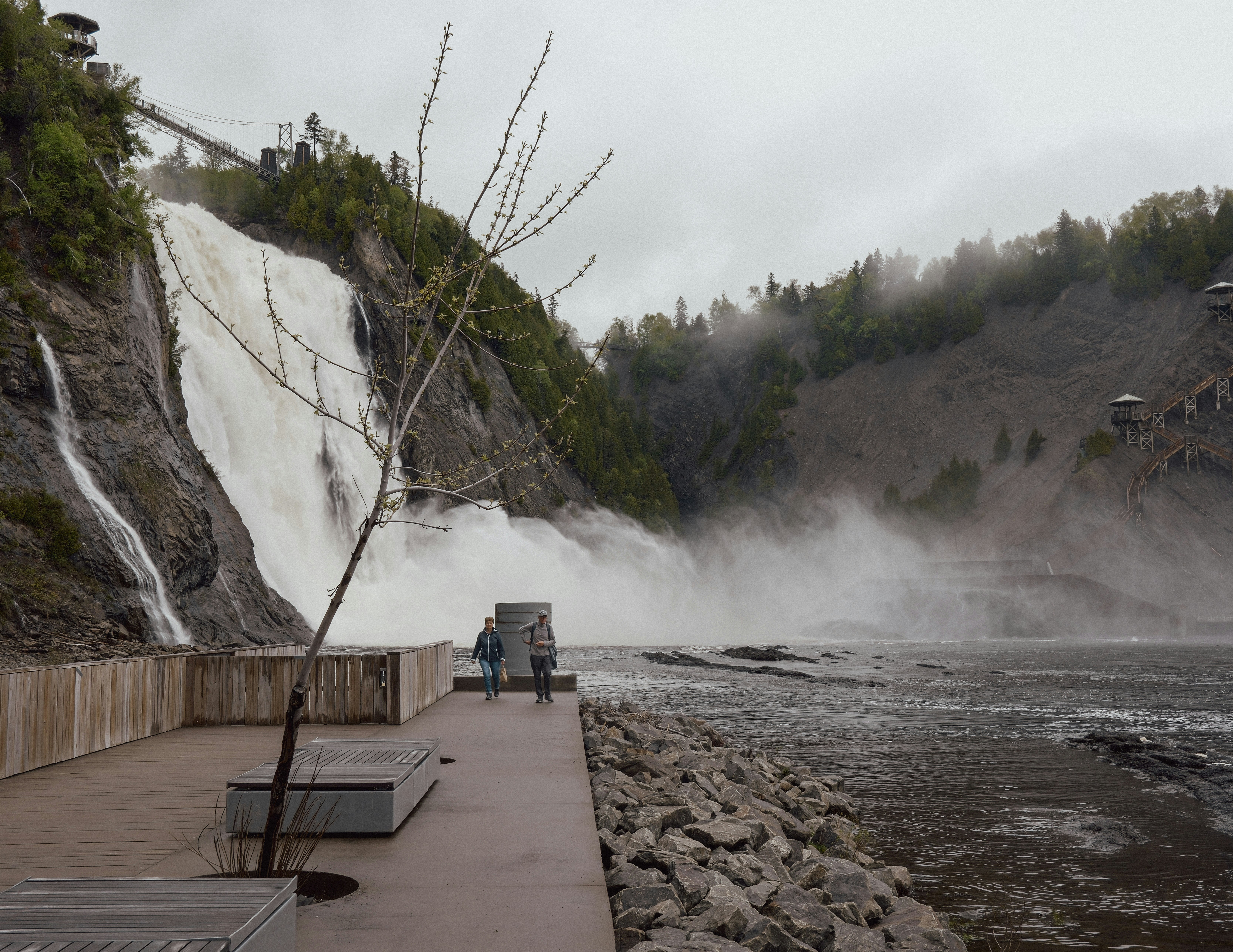 A low-angle vertical photograph captures two people walking along a concrete promenade right at the base of Montmorency Falls. A massive wall of rushing white water and heavy mist towers over the walkway on the left, creating a dramatic sense of scale. A rocky shoreline borders the churning river waters on the right under a cloudy sky. This photo was taken by Fujifilm xt5 with 23mm and no edit just fuji color, emphasizing the powerful, immersive art and crisp textures of the landscape.