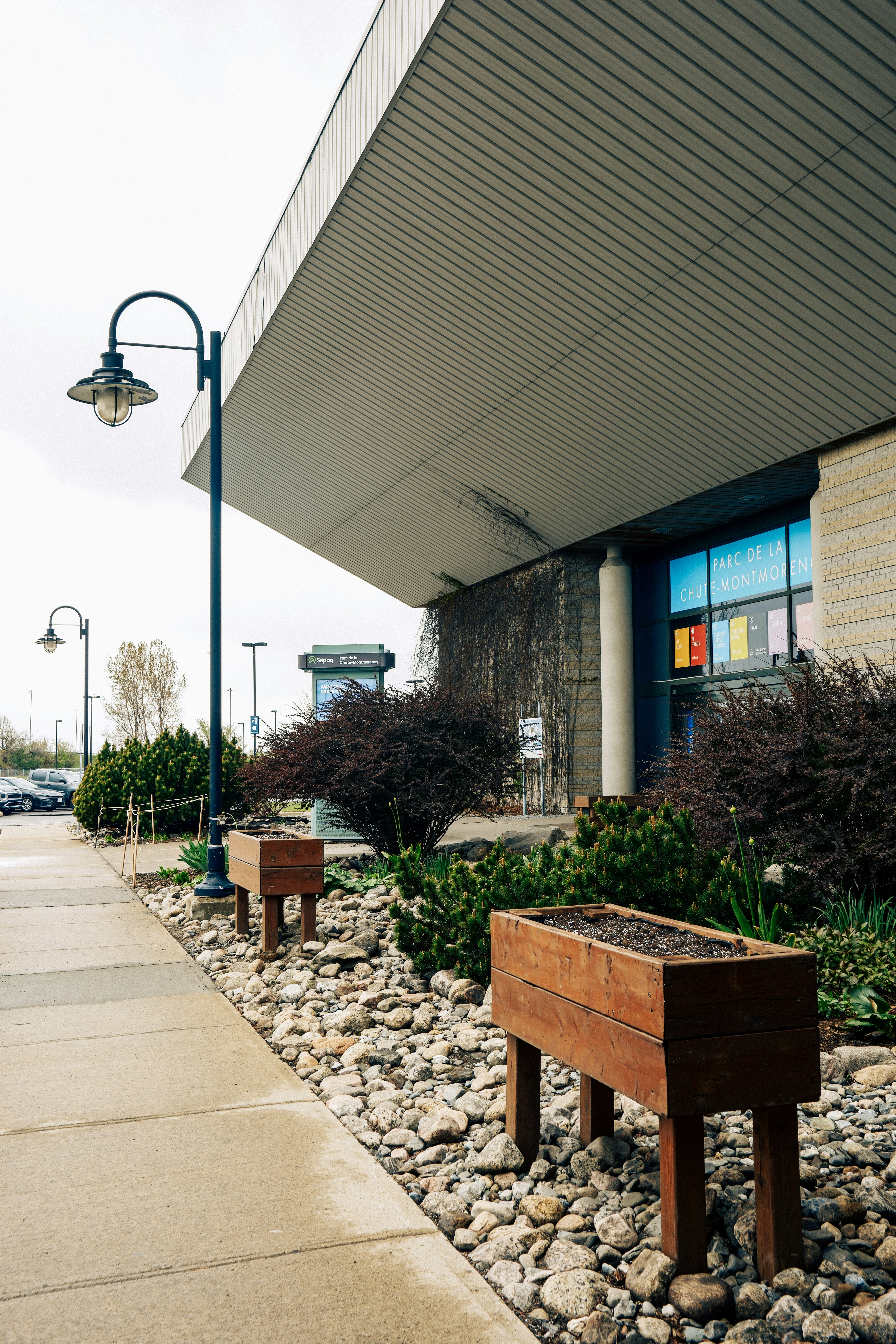 A vertical street-level photograph showcases the modern entrance of the Parc de la Chute-Montmorency visitor center. Wooden planter boxes filled with fresh soil sit on a rocky garden bed next to a smooth concrete sidewalk. A classic black lamppost frames the left side, while the building's large overhanging roof creates strong architectural lines against a bright sky. This photo was taken by Fujifilm xt5 with 23mm and no edit just fuji color, capturing sharp textures and clean, modern park design.