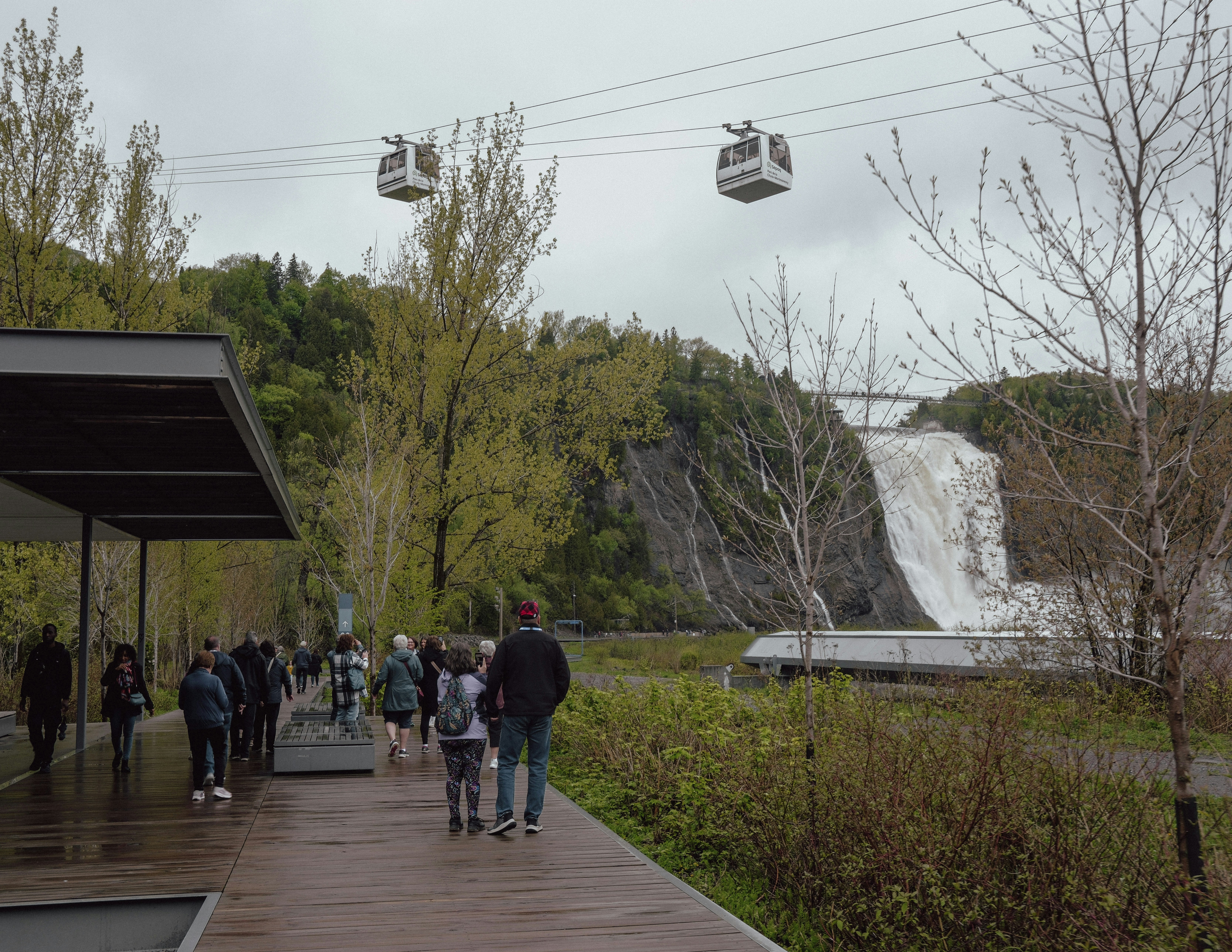 A wide, street-level photograph captures visitors strolling along a wet wooden boardwalk path toward Montmorency Falls. Overhead, two white cable cars glide along suspension wires against a pale sky. The massive rushing waterfall dominates the background, framed by lush green trees and a modern viewing pavilion on the left. This photo was taken by Fujifilm xt5 with 23mm and no edit just fuji color, perfectly preserving the authentic social atmosphere, natural movement, and beautiful landscape layout of this famous tourist park.
