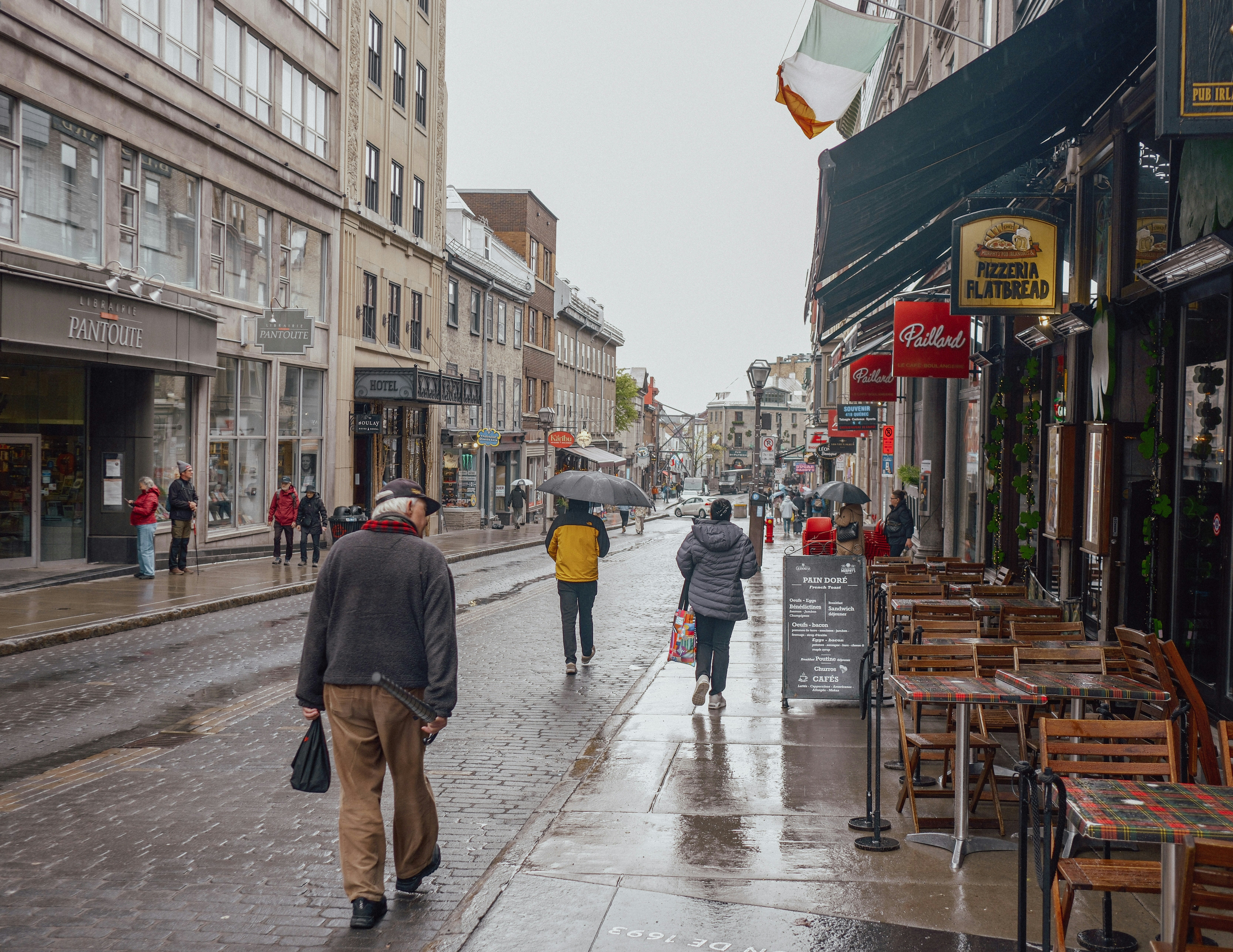 A wide horizontal photograph captures a rainy day on a historic cobblestone street in Old Quebec. Pedestrians walk along the wet pavement, some holding umbrellas to shield themselves from the drizzle. On the right, a charming restaurant patio features wooden tables and chairs set up under a dark green awning, next to a sign for a pizzeria. This photo was taken by Fujifilm xt5 with 23mm and no edit just fuji color, beautifully capturing the glossy reflections on the ground, historic storefronts, and cozy, rainy-day urban mood.