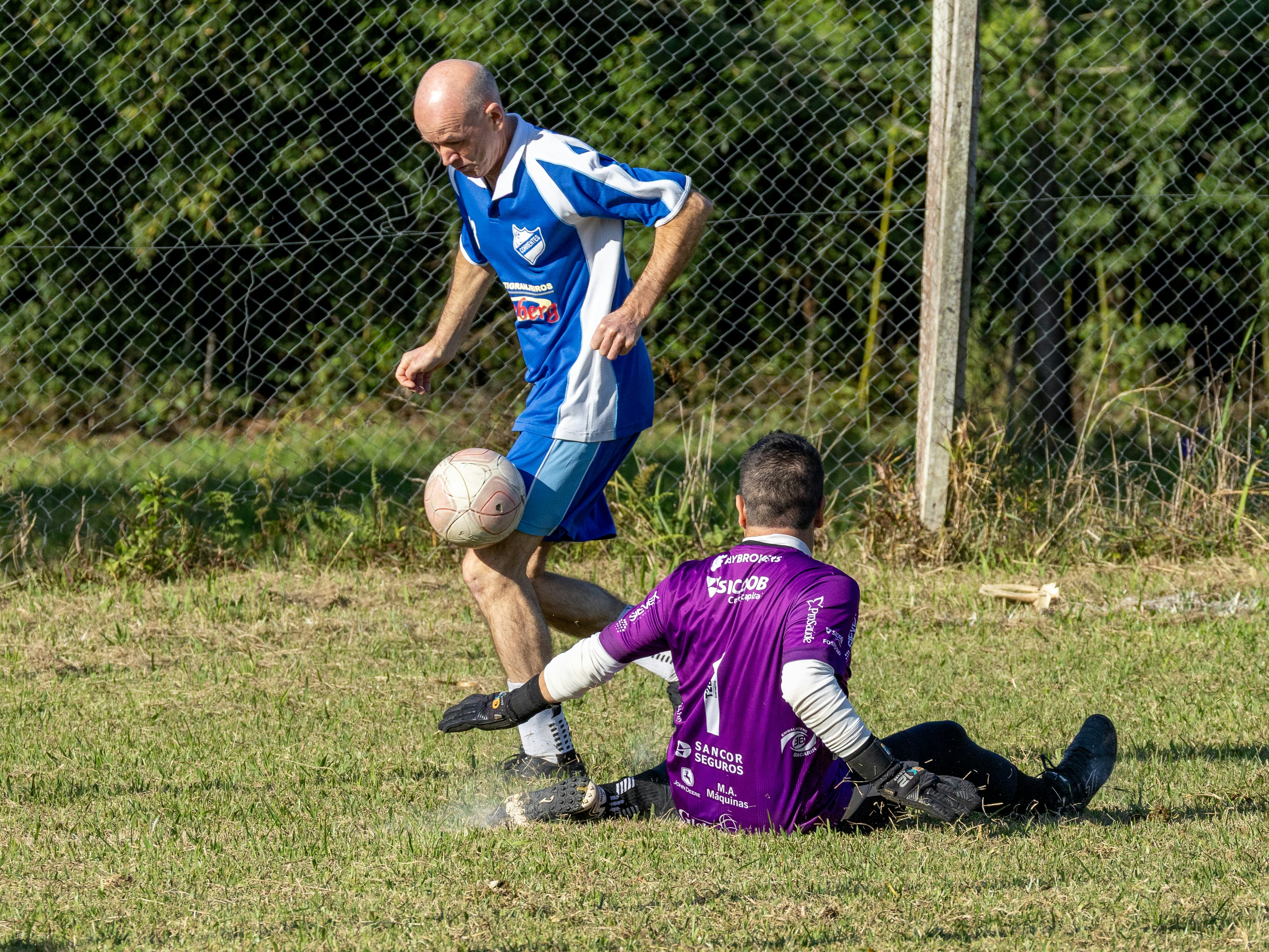 Soccer player in blue dribbles past a purple-clad goalkeeper.