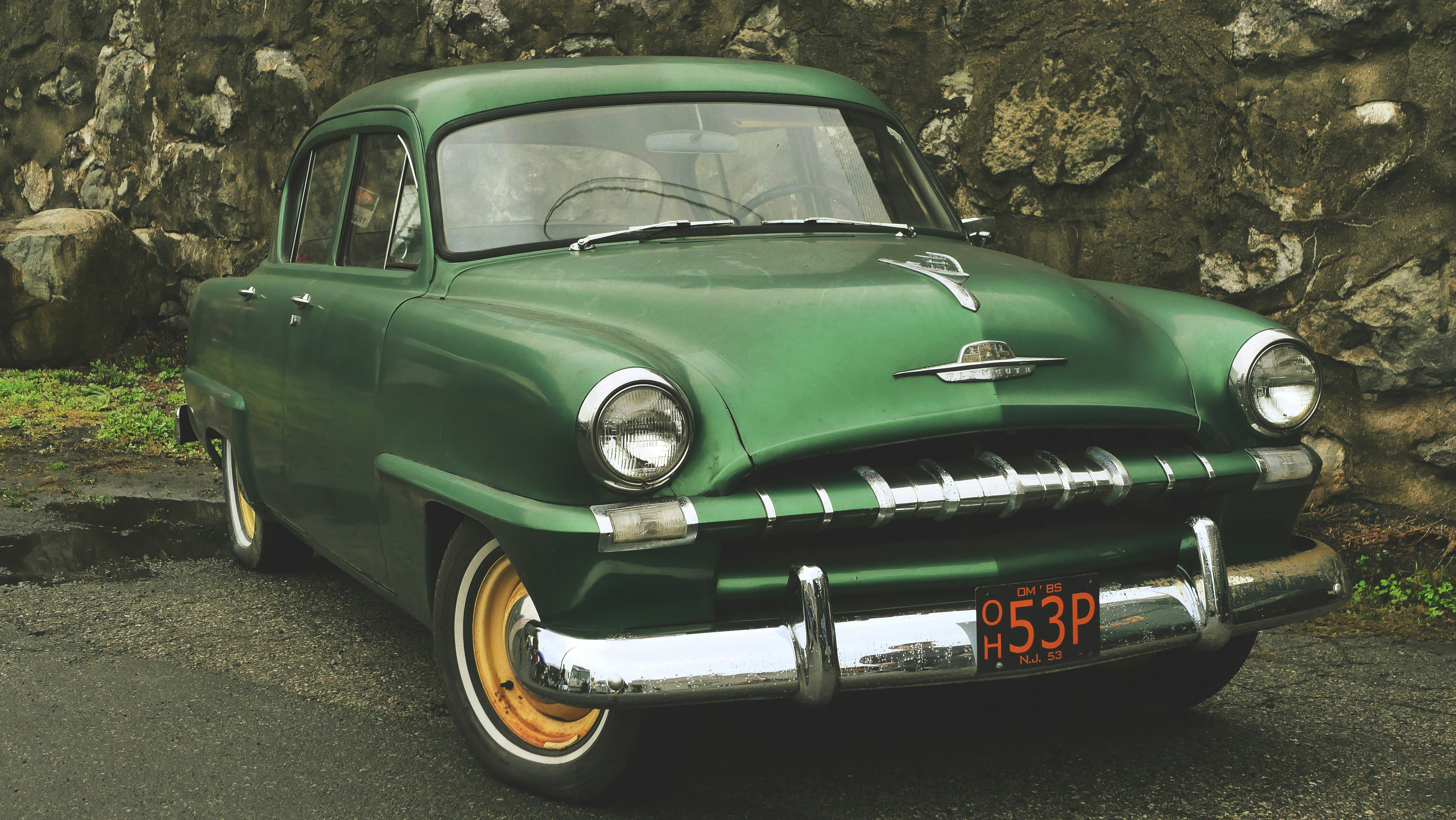 A vintage green Chevrolet sedan parked on a damp road by a rocky backdrop, showcasing chrome details and tan wheels. This is a photographic capture of a classic automobile, with attention to its nostalgic styling.