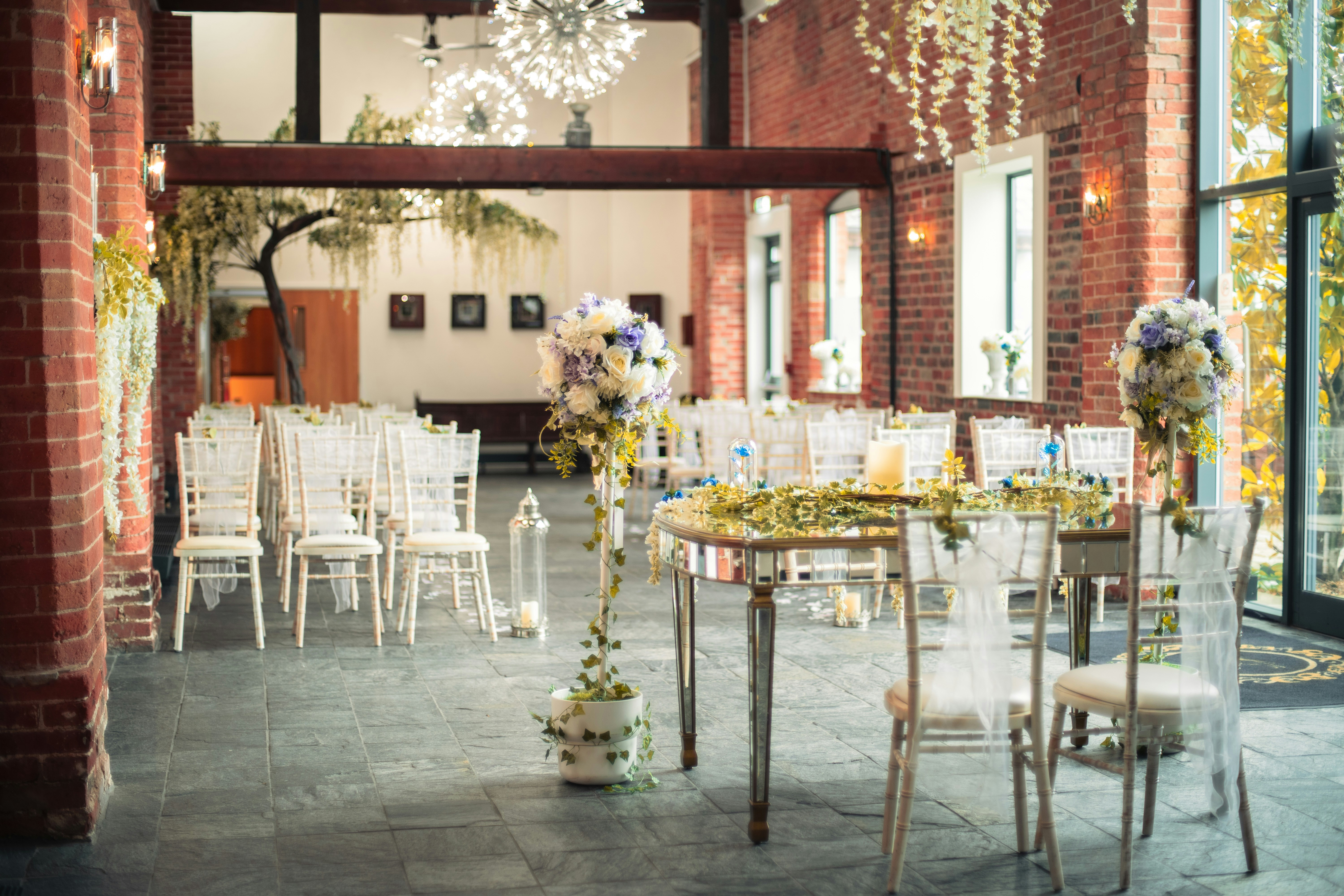 A beautifully styled wedding reception room at Goosedale, featuring exposed red brick walls, chiavari chairs, and towering floral centrepieces of white roses and purple blooms. Trailing wisteria and ivy cascade from the ceiling, while warm candlelight and natural light pour through floor-to-ceiling windows.