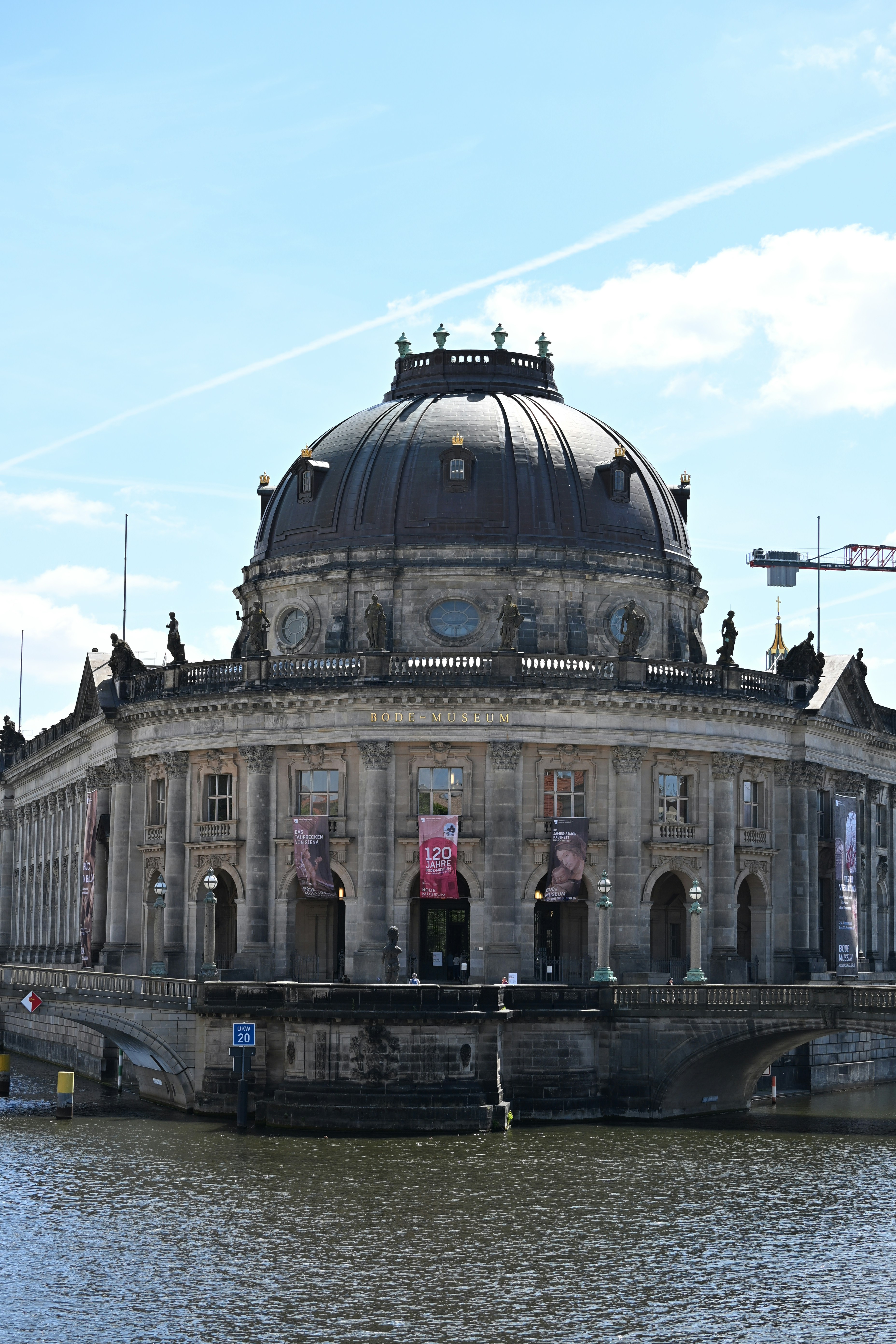 Baroque-style museum building sits on a riverbank with a large domed roof and arched entryways; banners hang from its façade, reflecting in calm water.