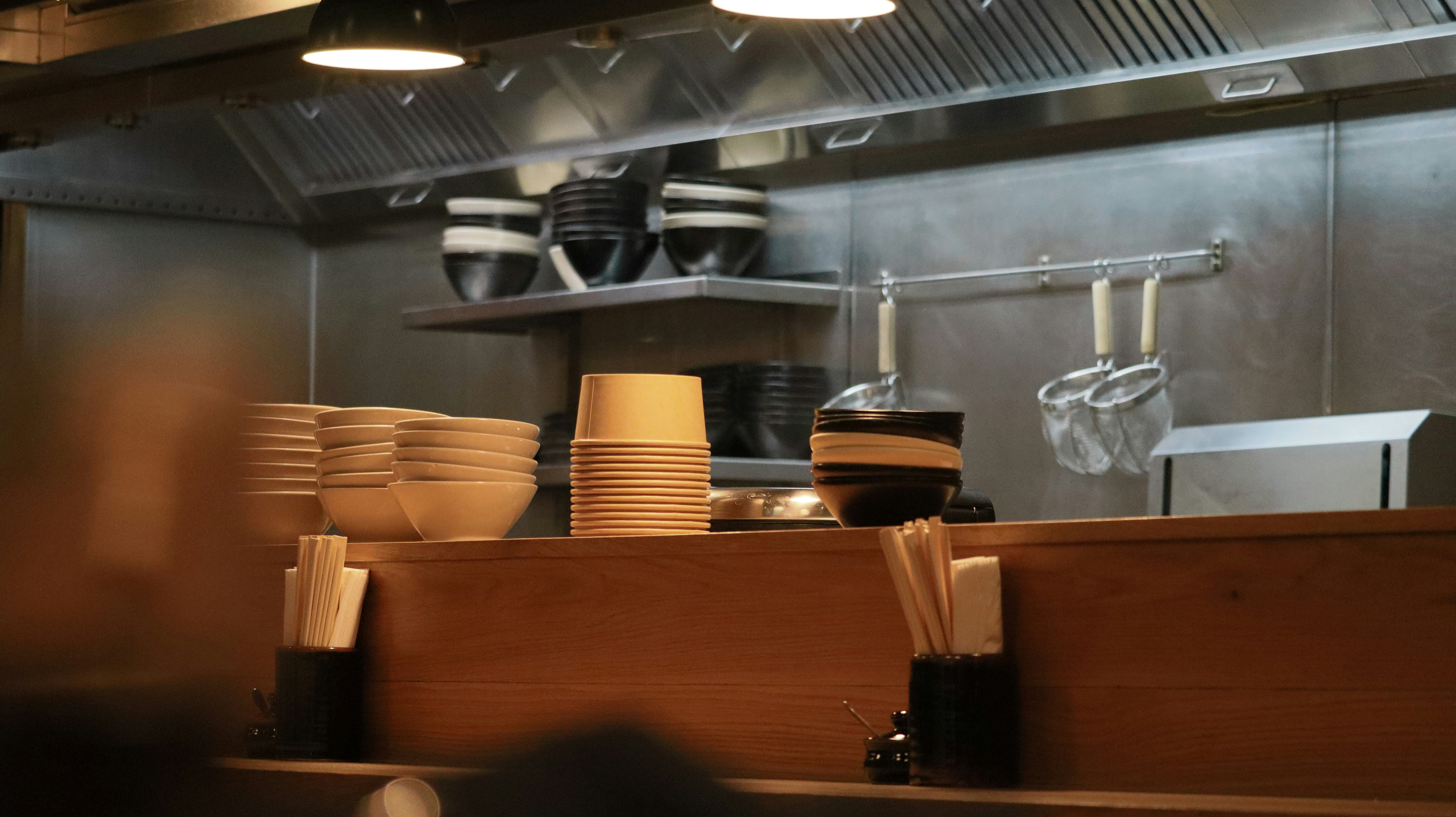 A blurred foreground shows a busy restaurant kitchen with stacked bowls and cups on a shelf behind a wooden counter; the scene emphasizes the behind-the-scenes prep area rather than a specific person.
