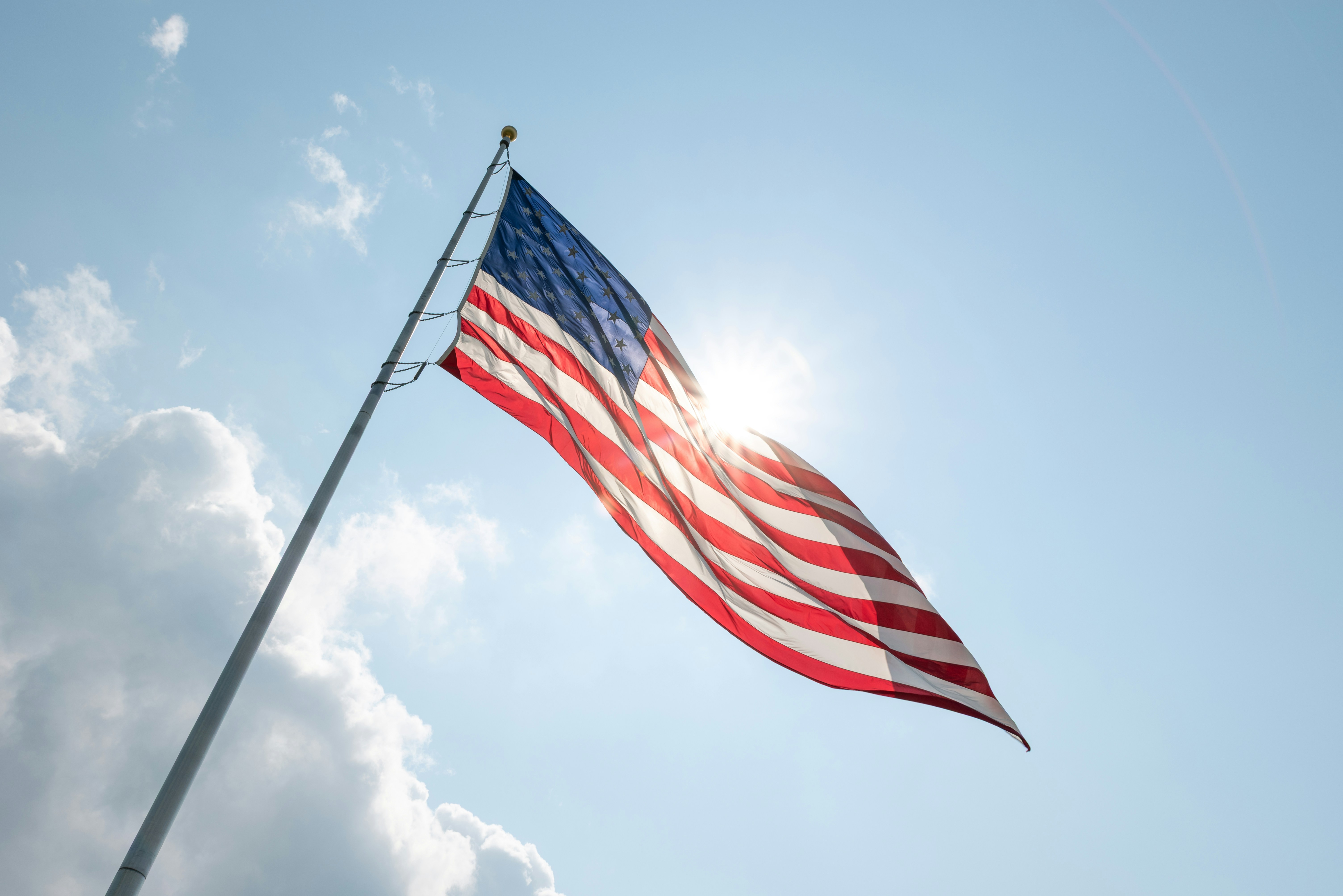 A striking backlit American flag glows as brilliant sunlight shines directly through the fabric from behind, illuminating the red, white, and blue with a luminous, almost ethereal warmth. The sun bursts through just behind the star field of the canton, creating a radiant halo effect that gives the image an uplifting, almost spiritual quality. The diagonal flagpole rises from the lower left while white cumulus clouds frame the left side, completing a powerful and emotionally resonant composition. The backlighting creates a translucent quality in the stripes that sets this image apart from any standard flag photo. Perfect for inspiration, hope, freedom, patriotism, national pride, Memorial Day, Veterans Day, Fourth of July, or powerful emotional Americana concepts.