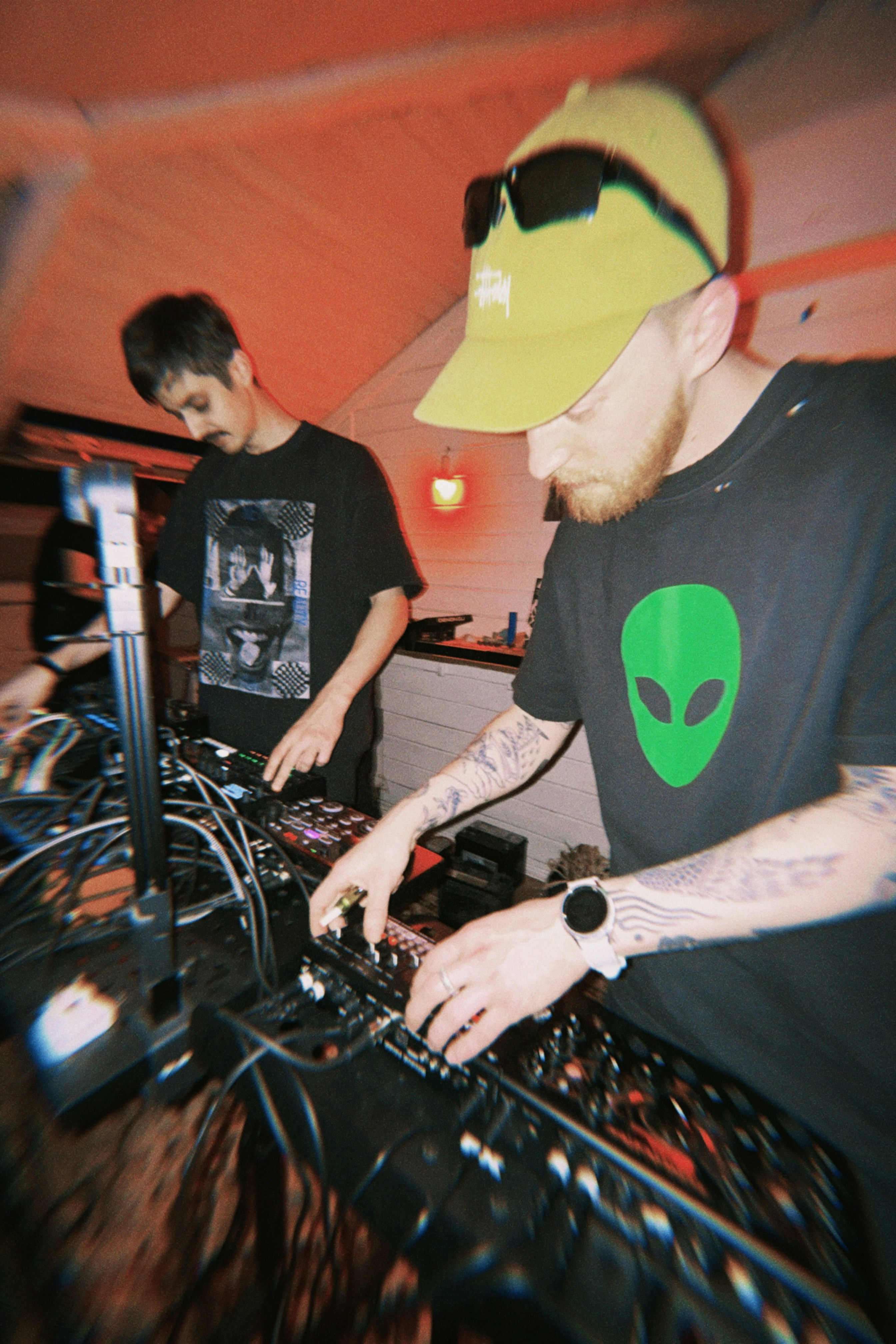 Men operating dj equipment in a dimly lit, reddish venue.