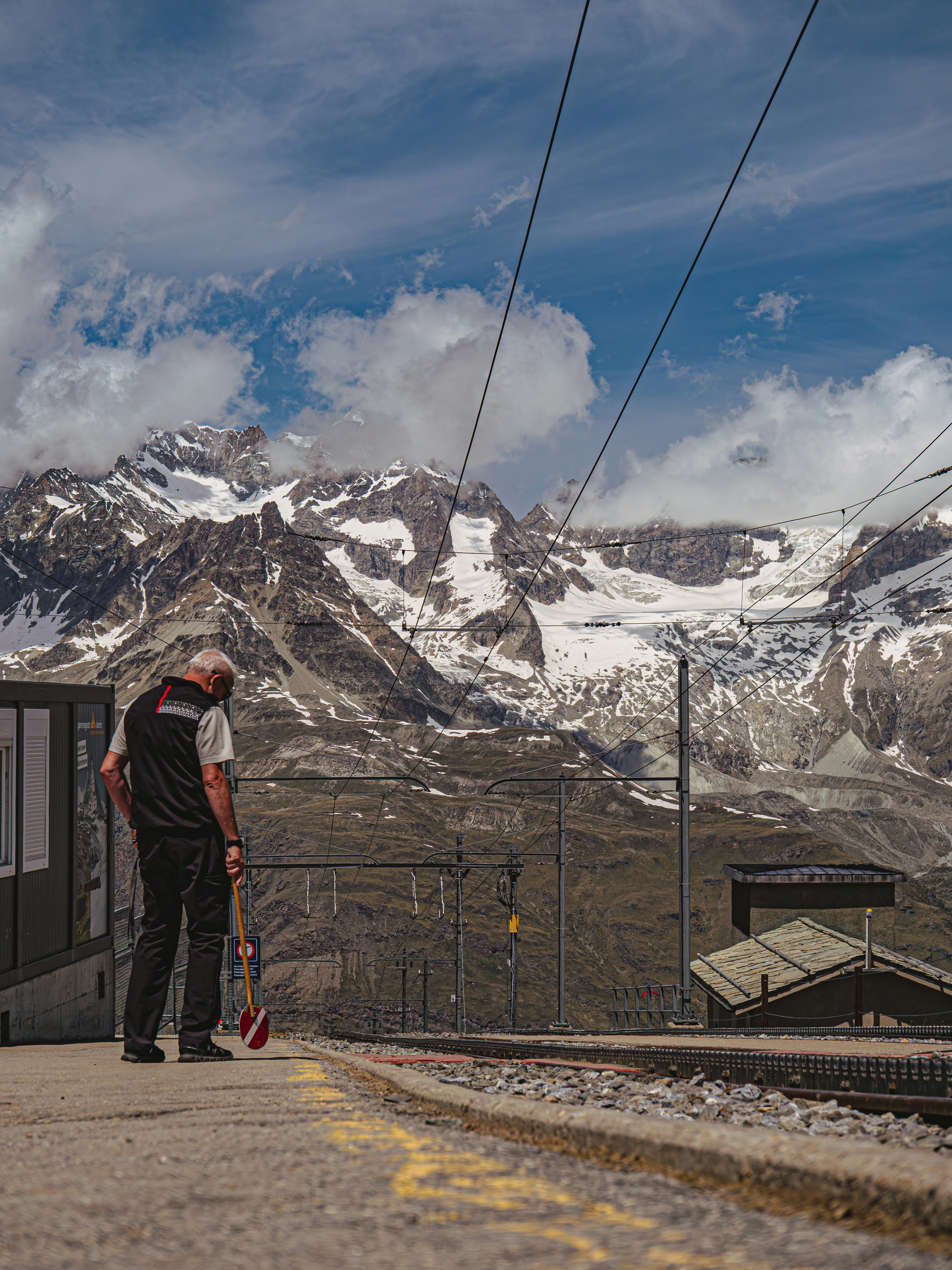 Railway worker holds signal paddle by tracks, snowy mountains beyond.