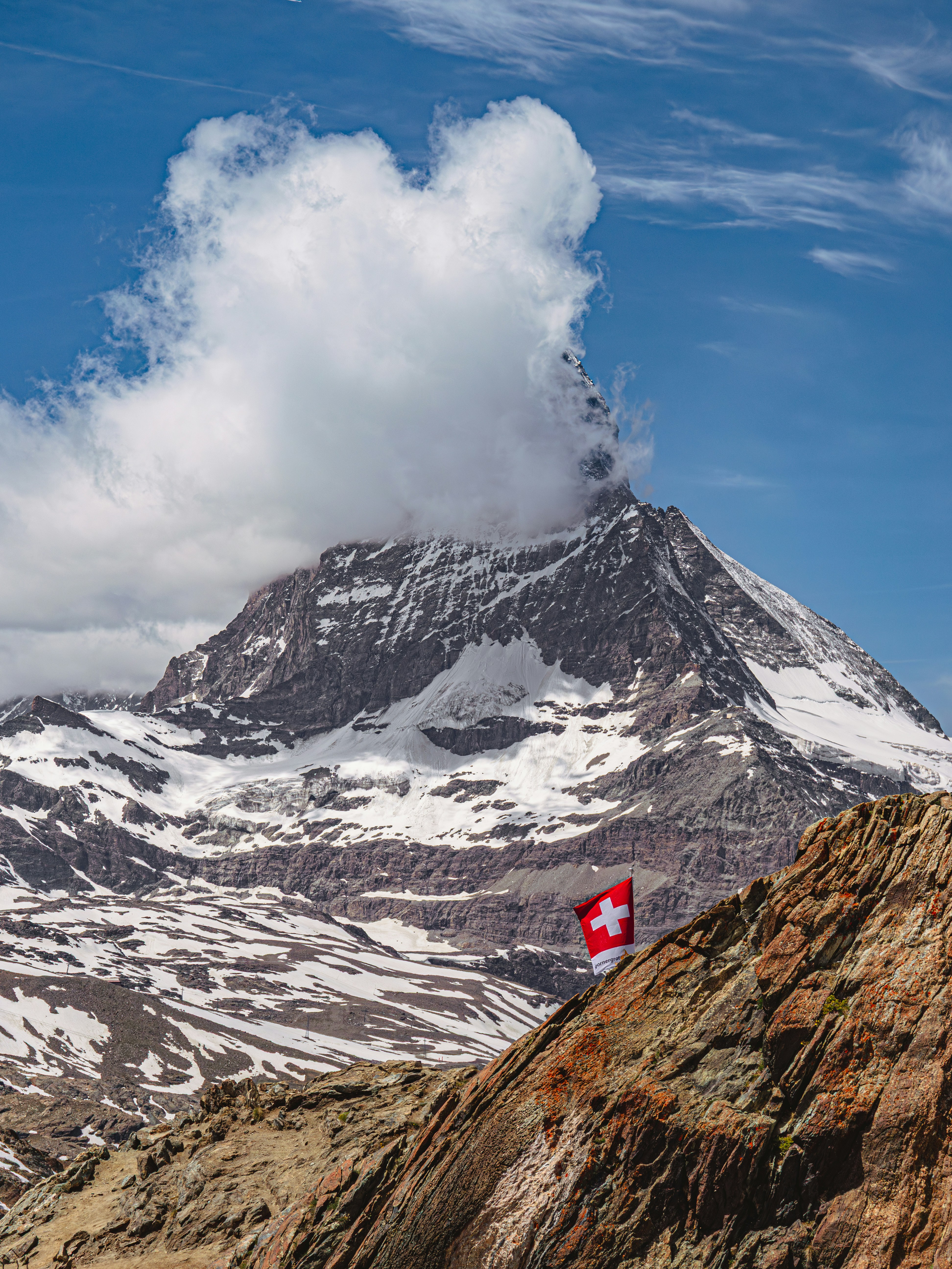 Swiss Flag on Rugged Peak Beneath a Crowned Cloud