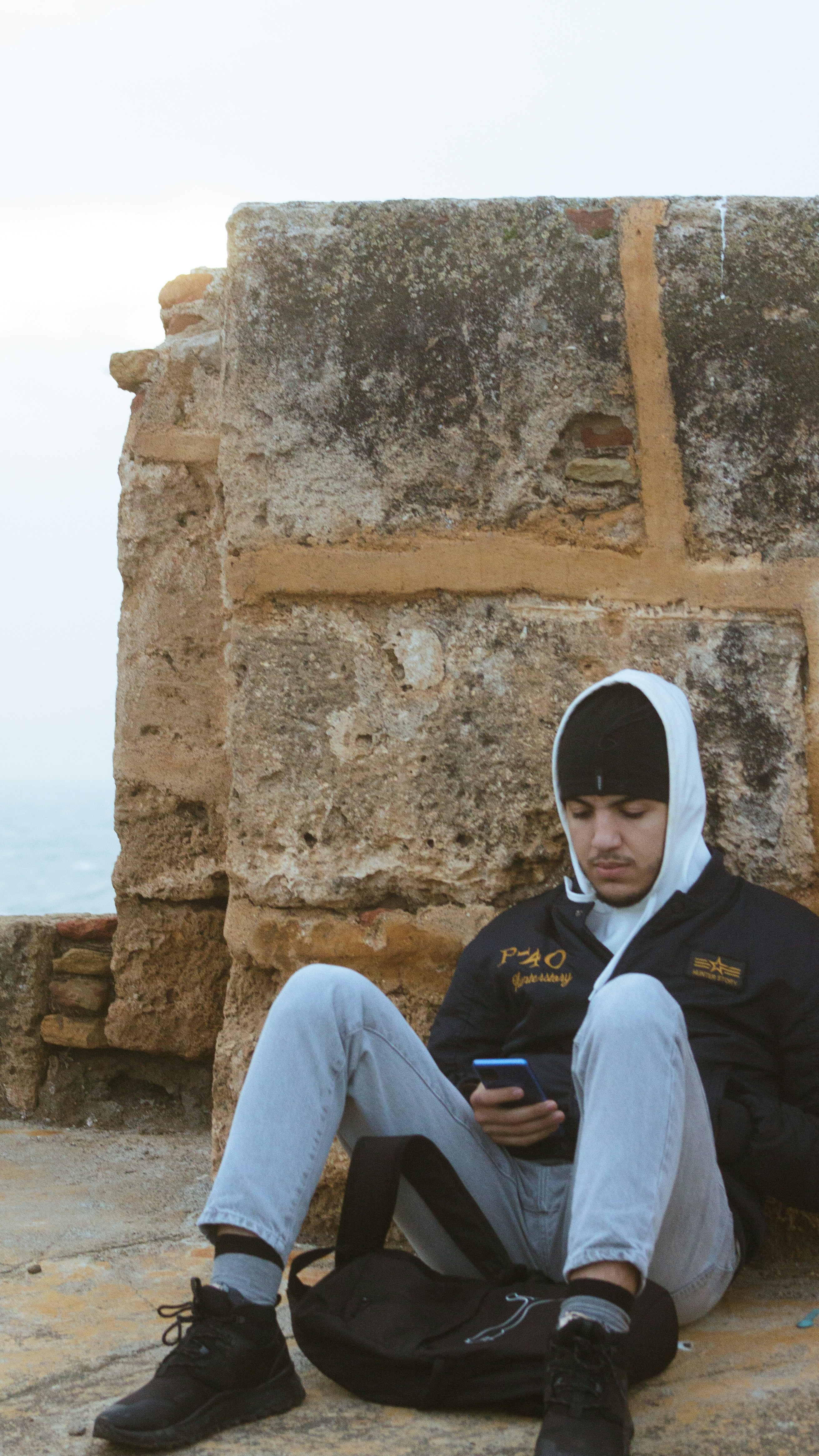 Teenager in a hoodie leaning against a stone wall, absorbed in a smartphone