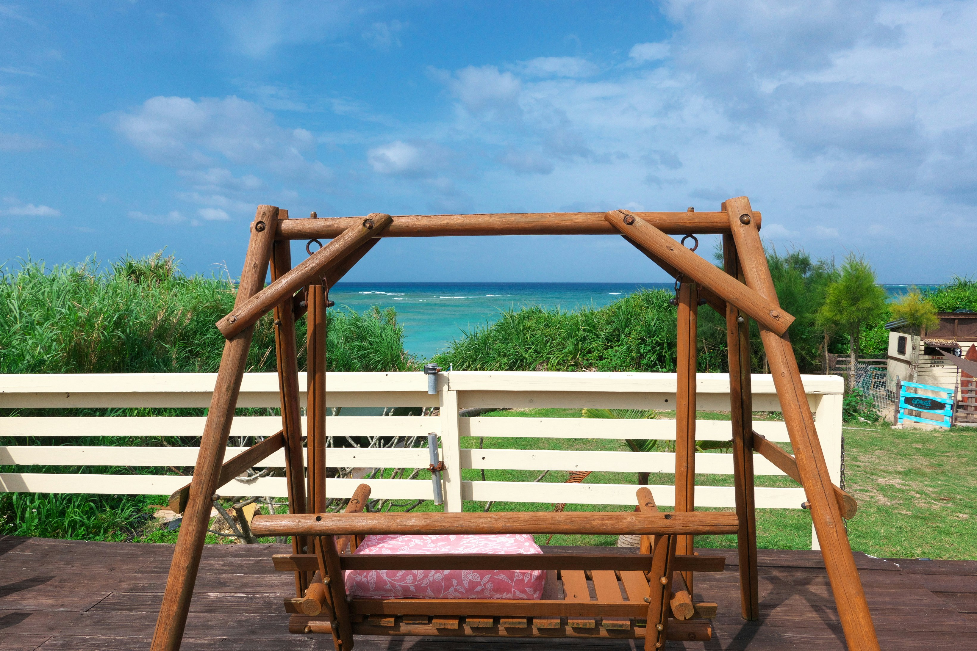 A wooden swing frame sits on a deck facing a coastal landscape with sea, dunes, and greenery in the background.