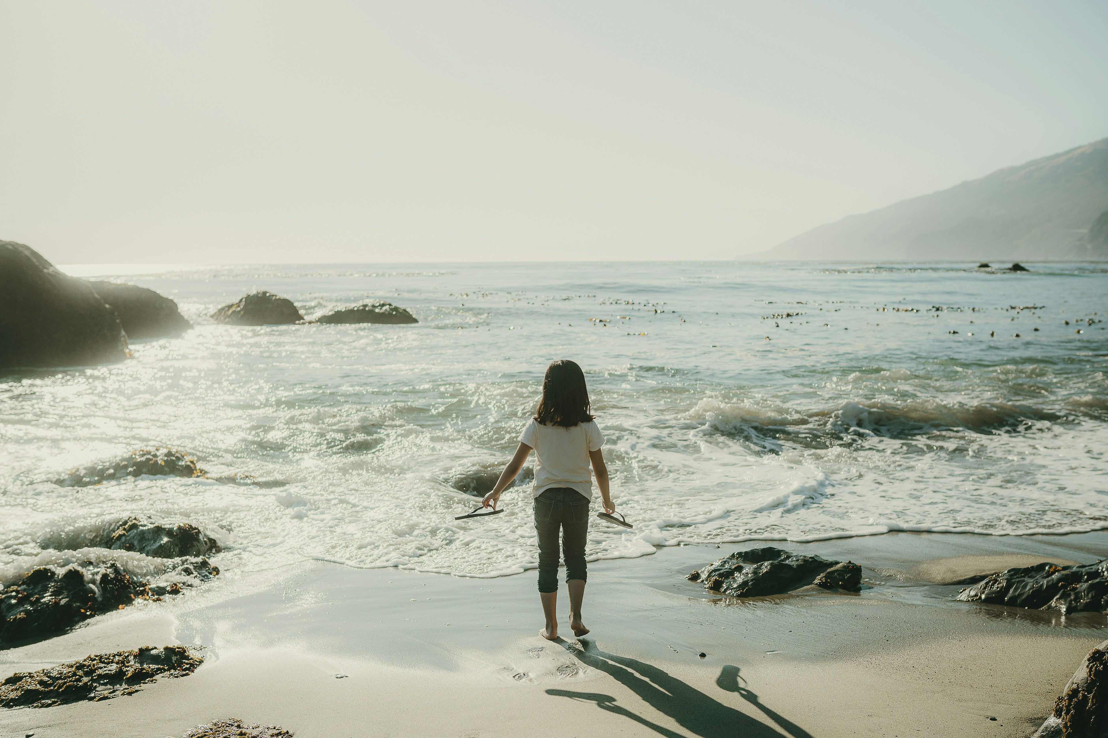 Sunlit beach moment with a girl standing at the shoreline, waves rolling in and soft golden light over the ocean.