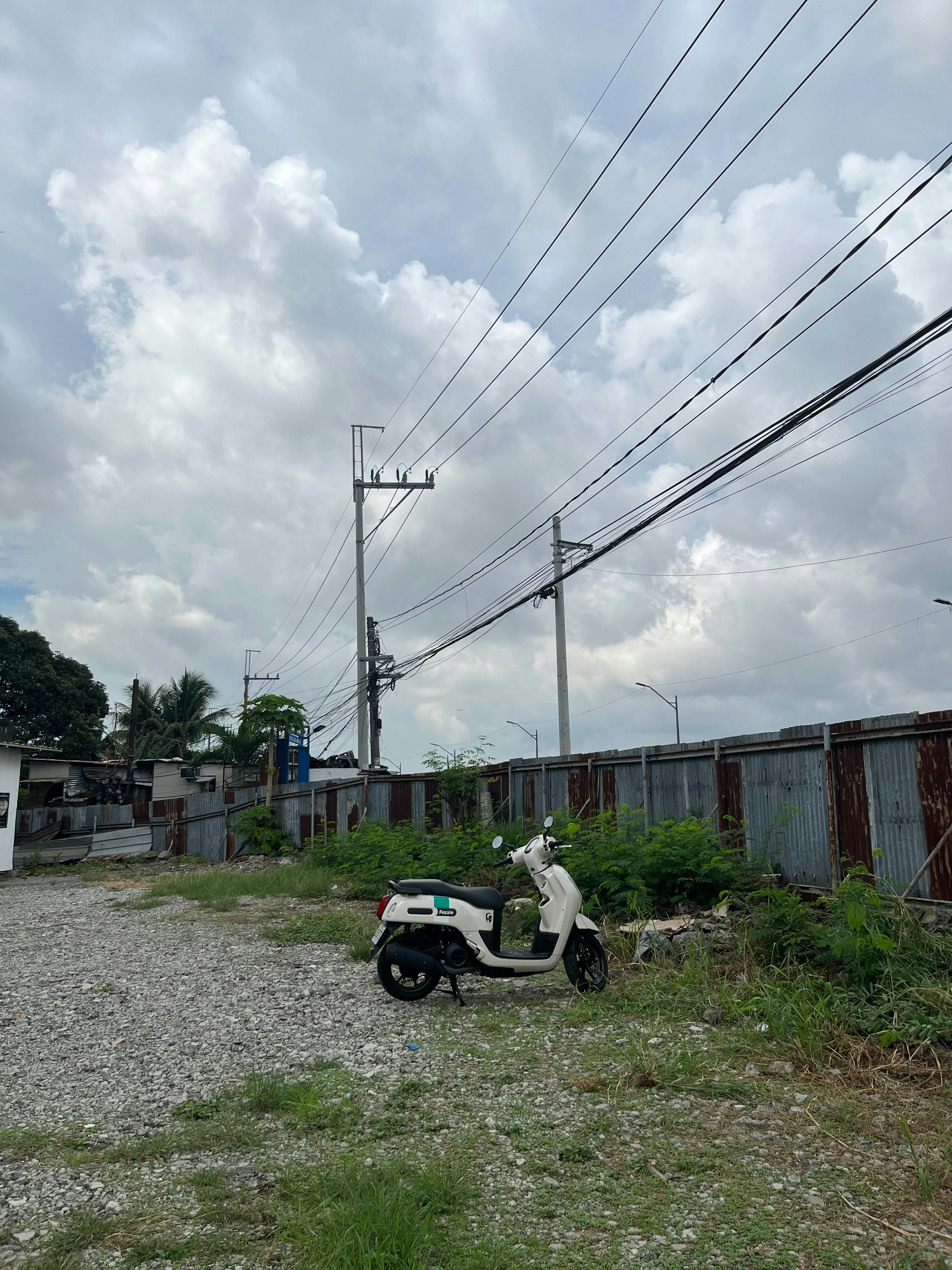 White scooter parked on a gravel lot beside a weathered fence with utility poles and power lines under a cloudy sky.