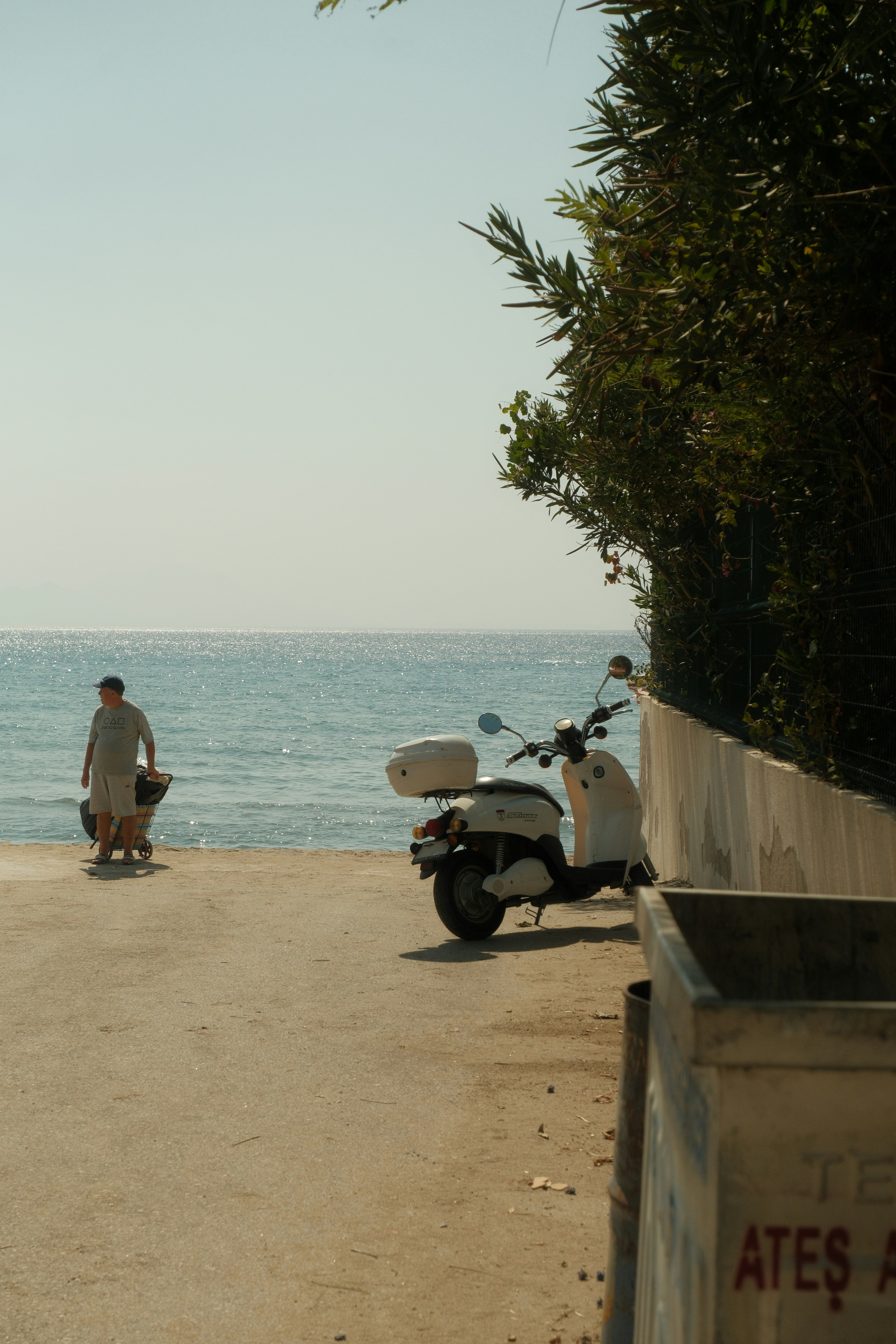 A quiet coastal pathway leading to the sea, with a white scooter parked beside a wall covered in greenery, while a lone man stands near the shoreline under soft midday light.