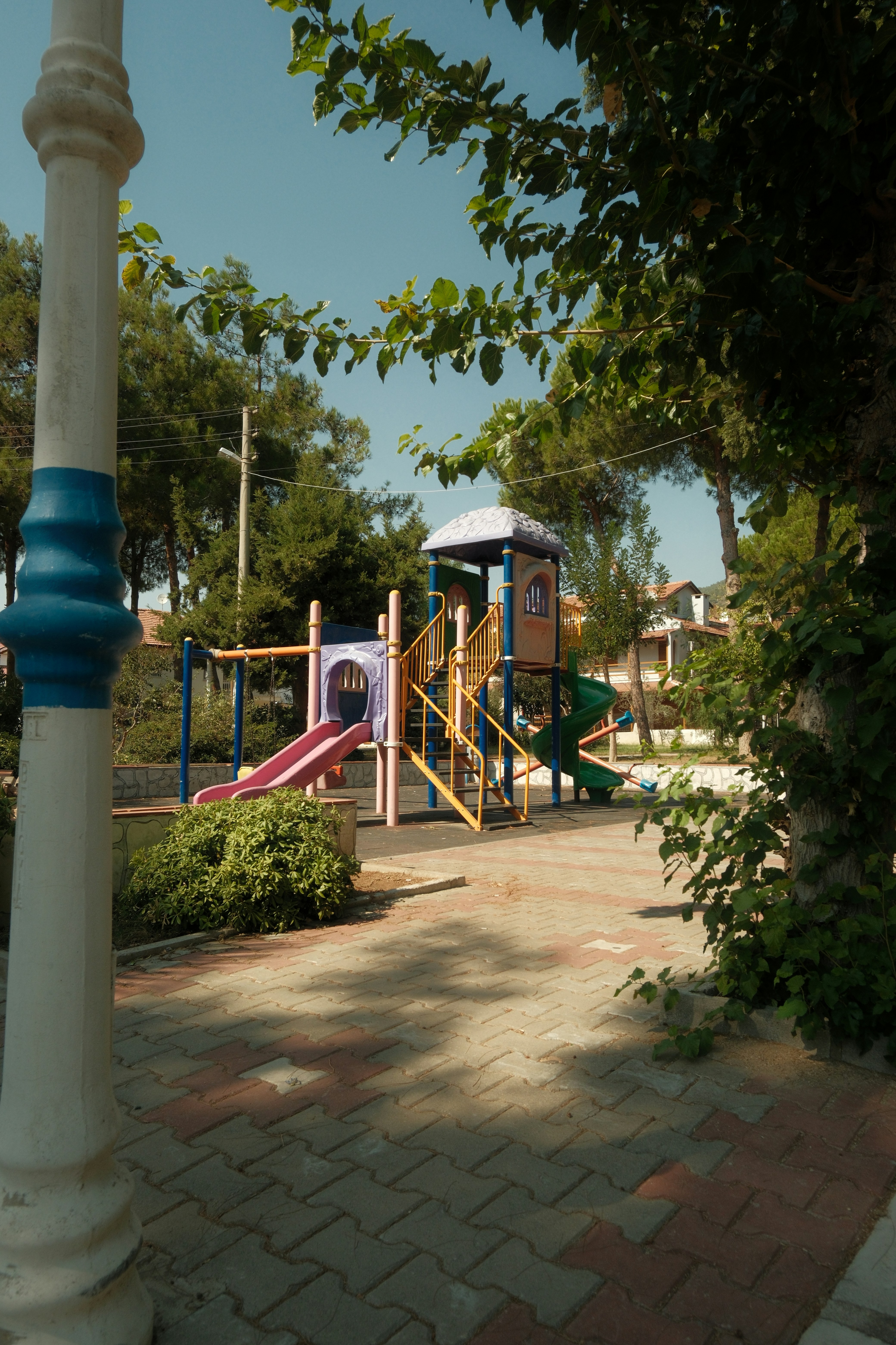 A colorful children's playground with slides and climbing structure sits empty on a brick-paved square, framed by pine and fig trees under a clear summer sky. The scene is observed through overhanging branches and a painted street lamp, giving the composition a candid, unhurried quality.