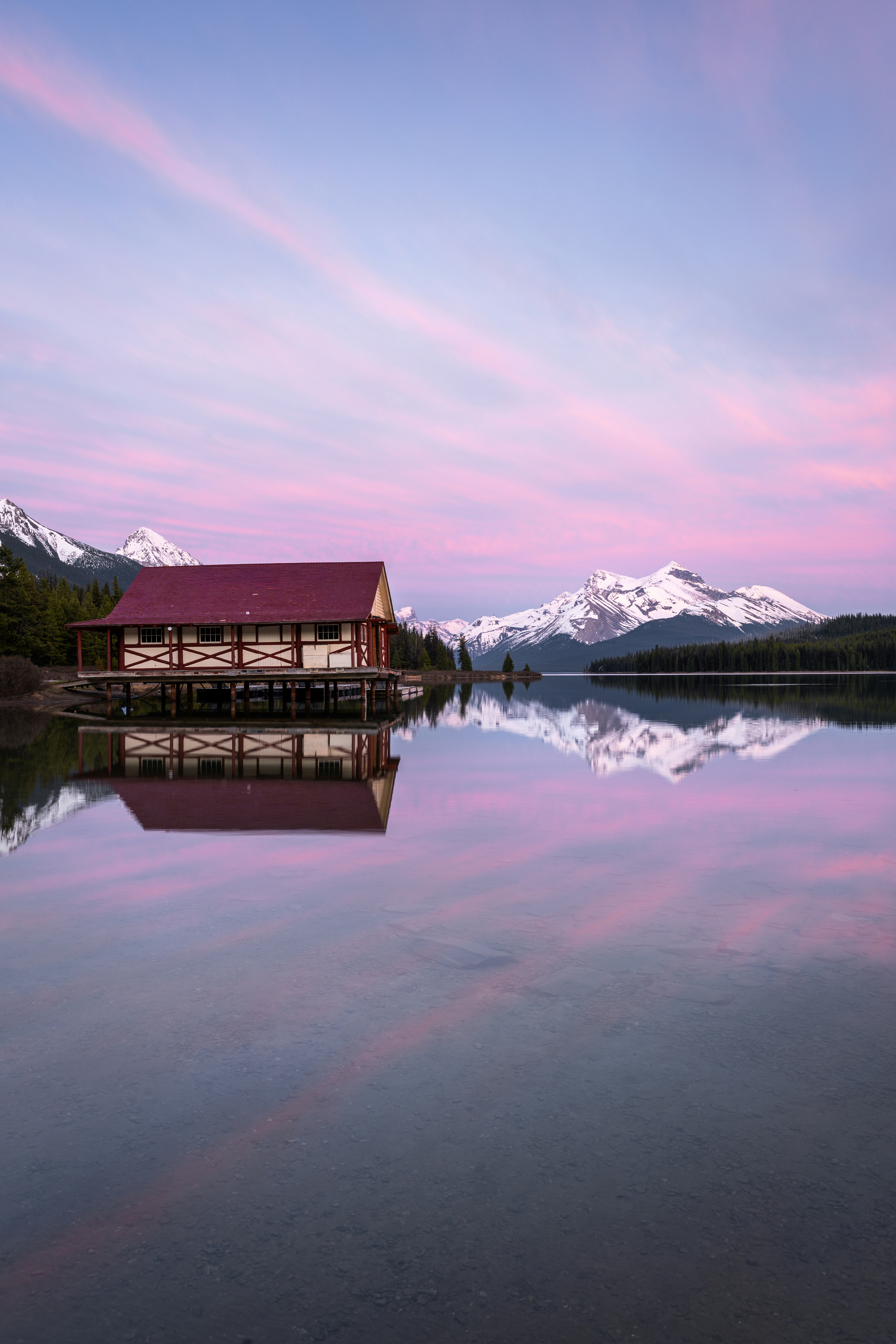 Serene lake with boathouse, snow-capped mountains, and pink sunset sky.