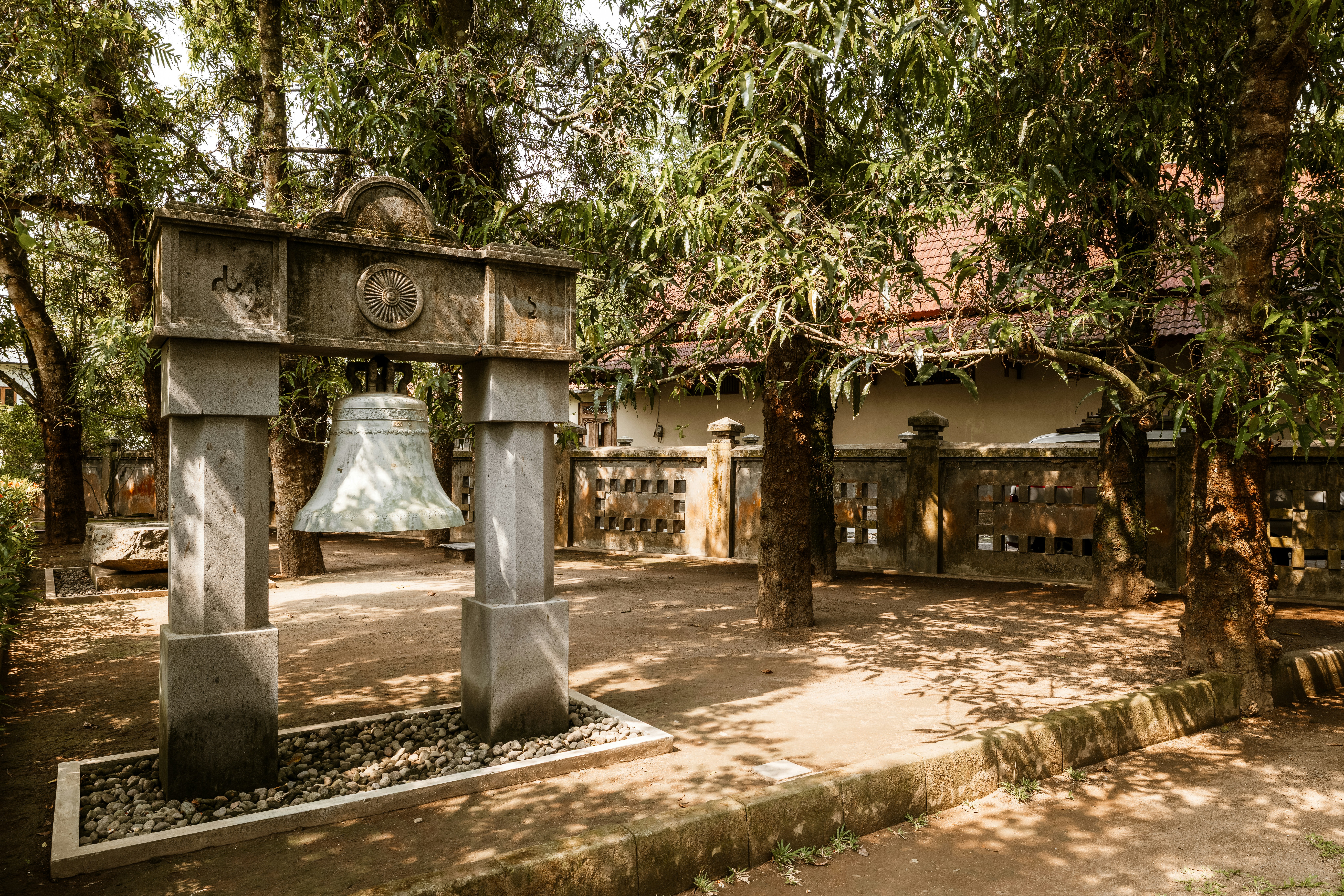 A large bell hangs within a stone archway amidst trees.