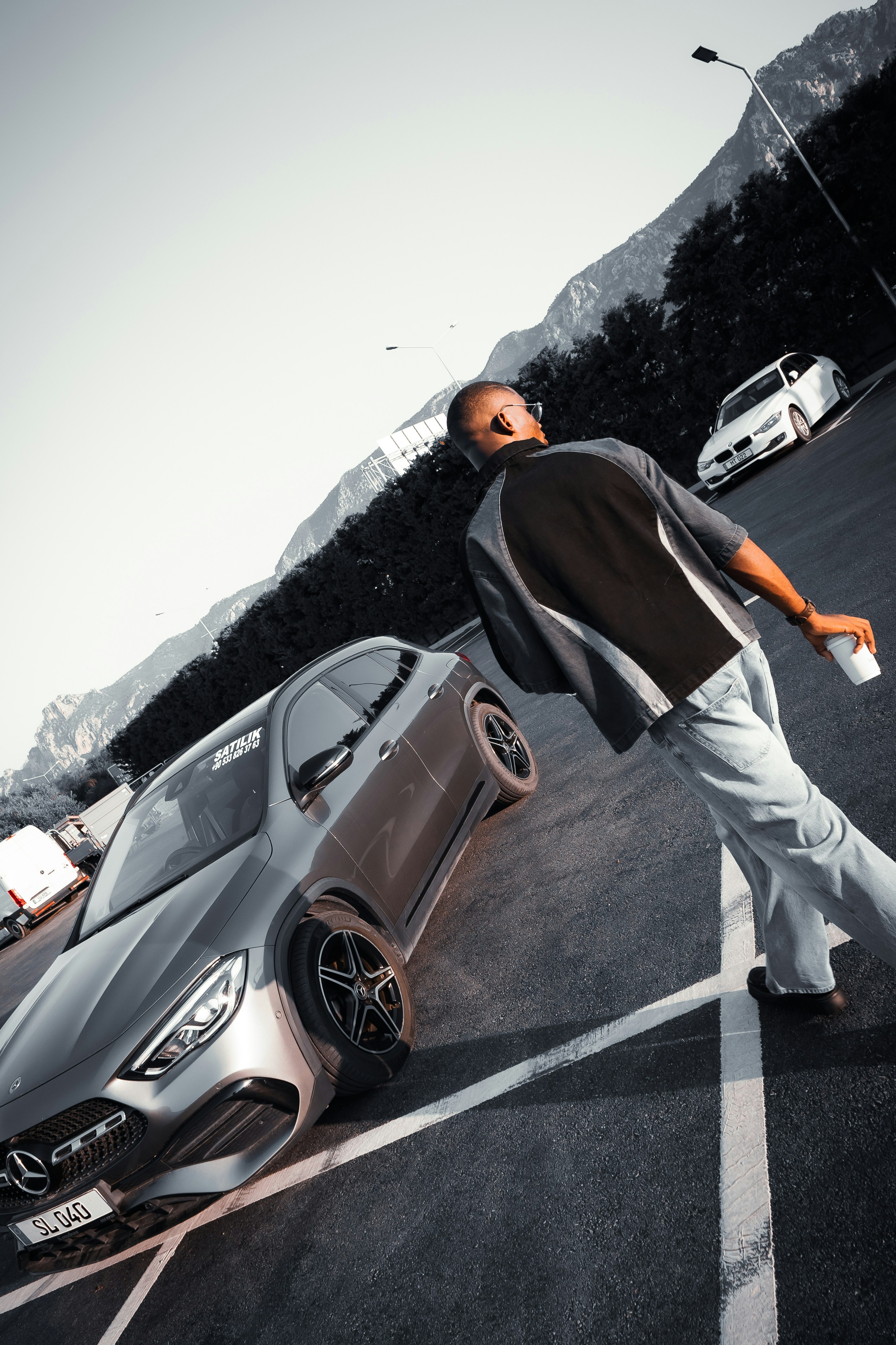 Leaned-Forward Street Scene: A man with beverage strides past a silver sportscar under a mountain pass