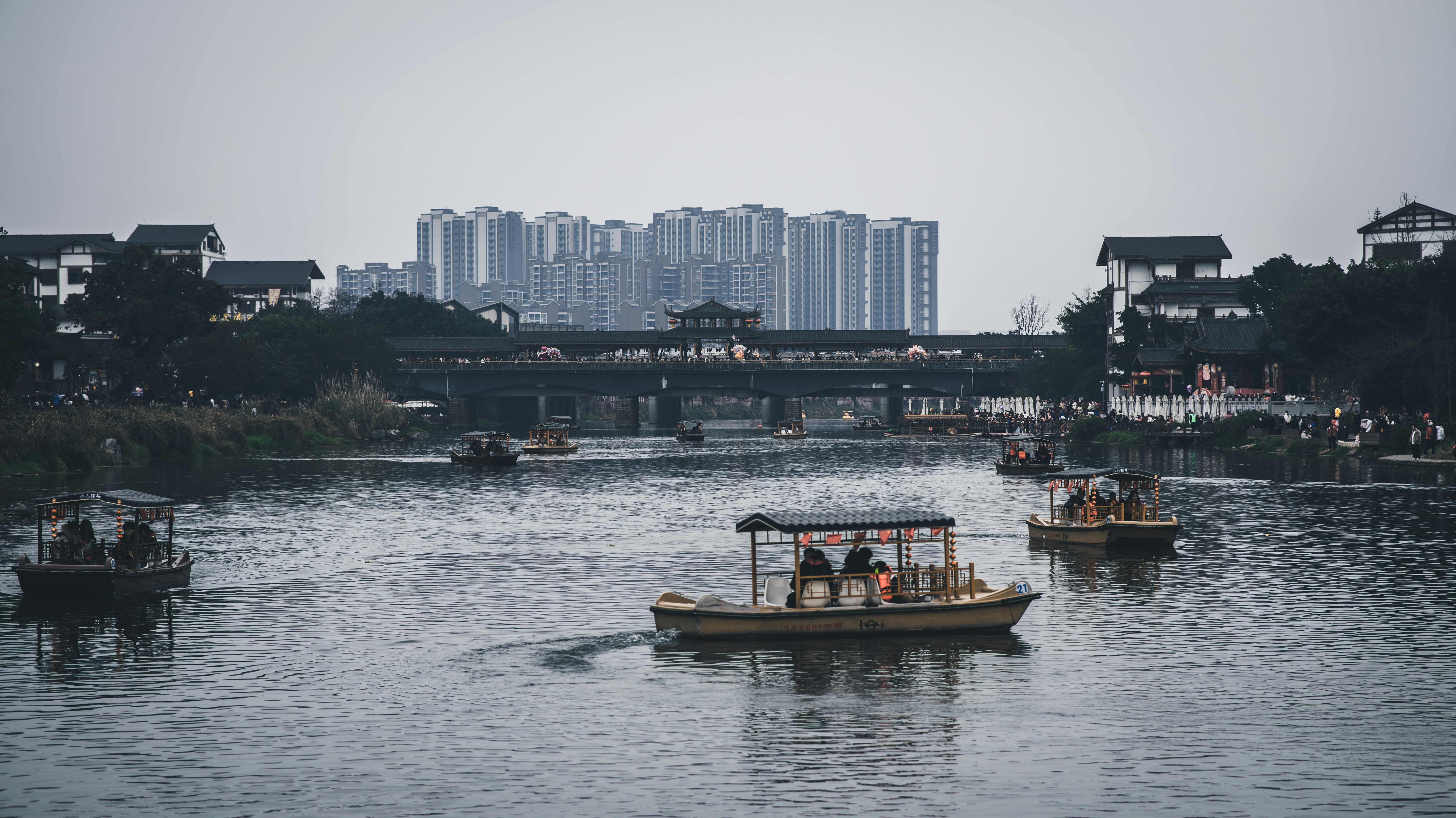 Calm river scene with small wooden boats and a distant cityscape; the composition centers on a boat in the foreground with a misty horizon and traditional structures along the banks.