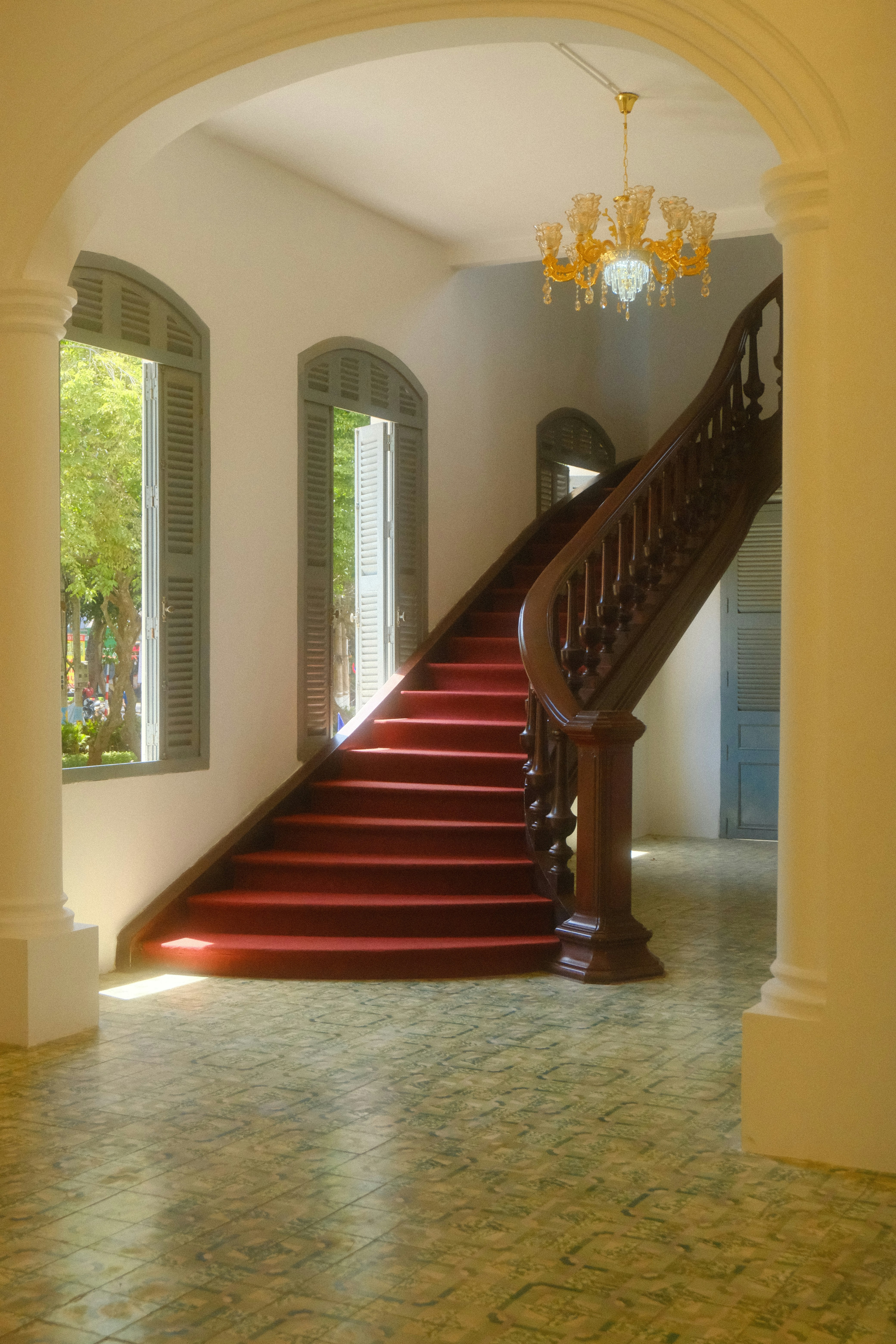 Ornate wooden staircase with red carpet and chandelier.