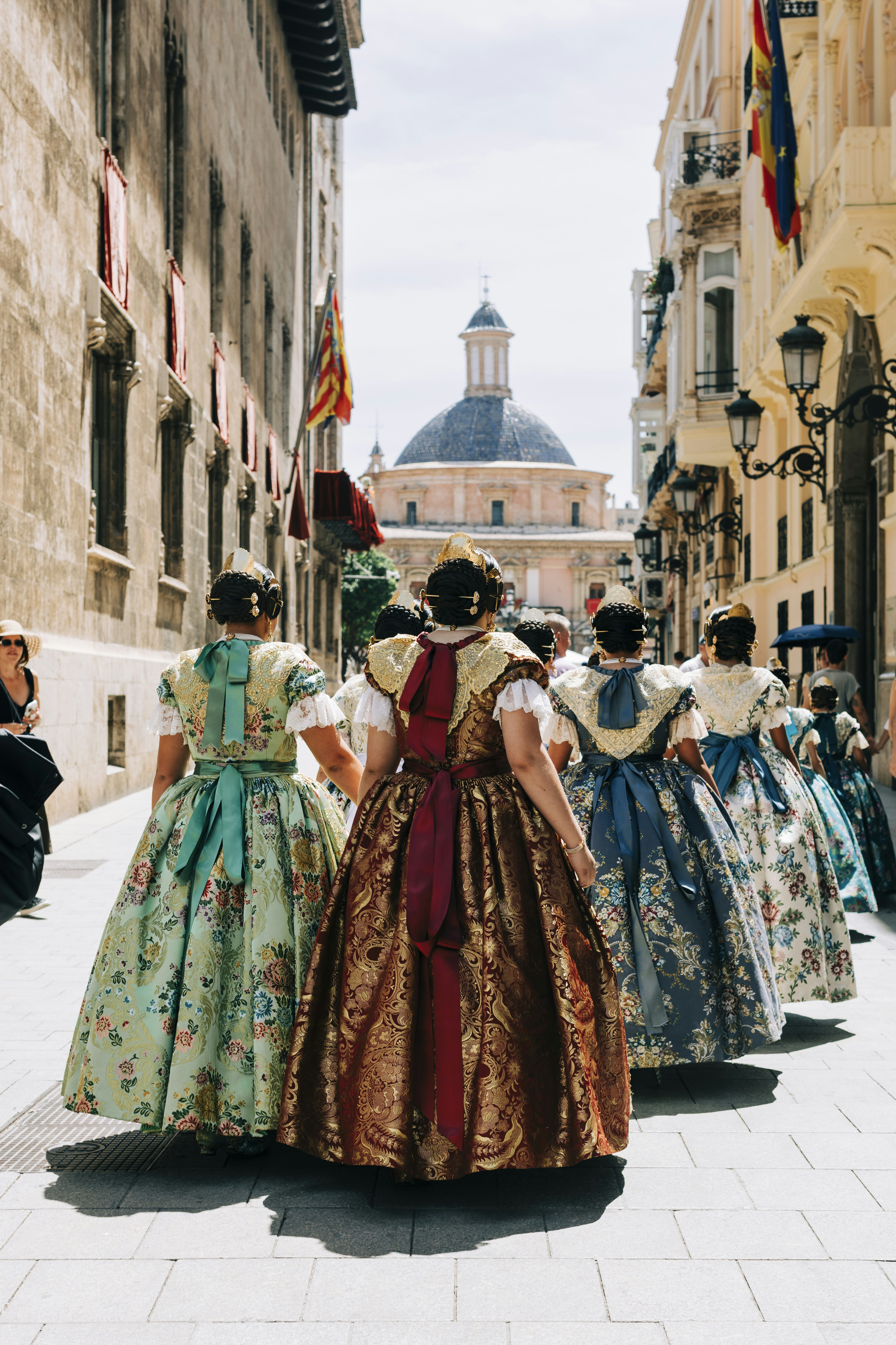 Procession in Full Dress: Victorian-inspired gowns march down a sunlit Spanish street toward a domed landmark