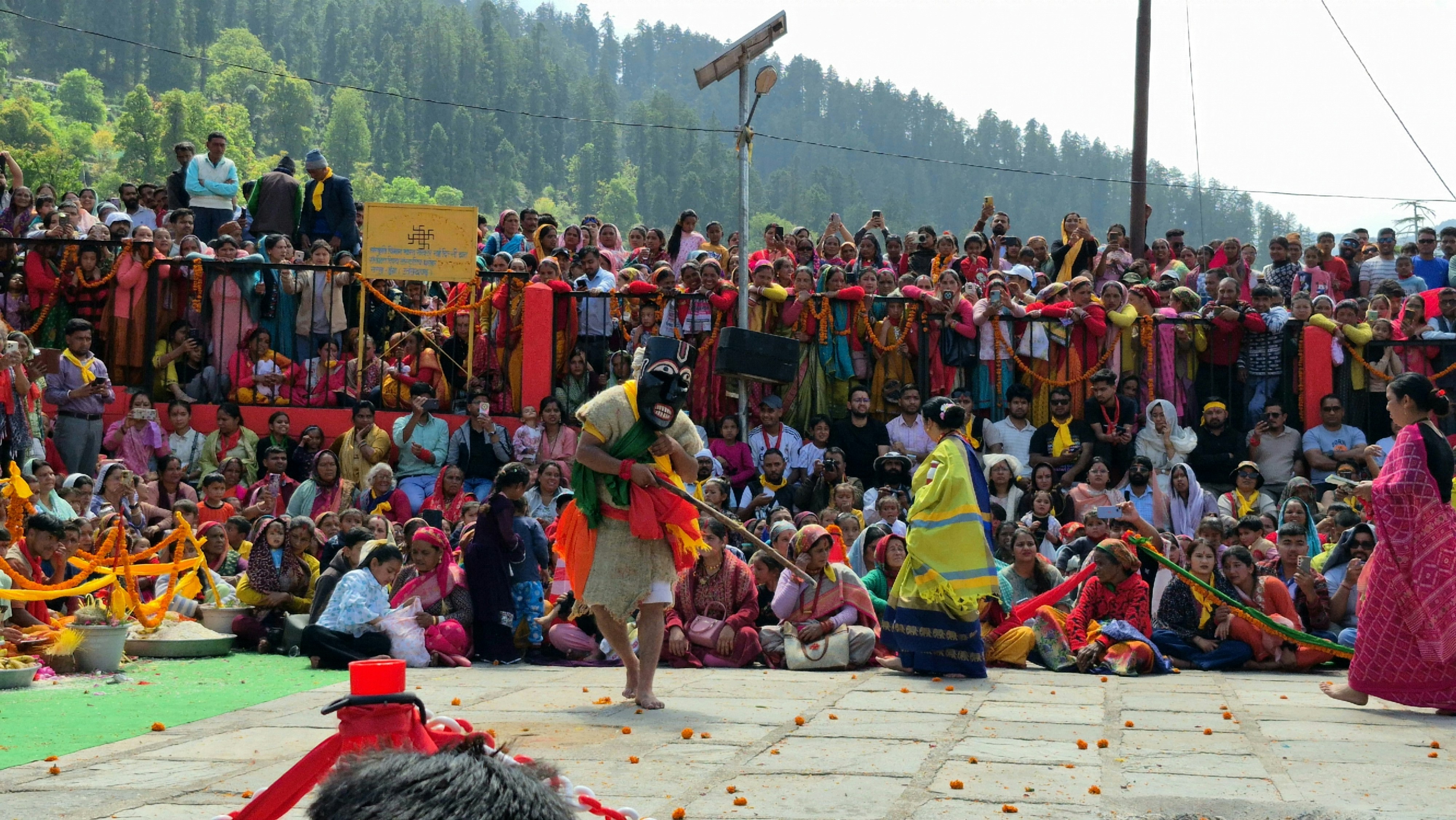 Ritual Theater Movement at UNESCO Ramman Mela, Uttarakhand. A captivating wide-angle shot capturing the lively interaction between traditional masked performers and dancers during the ancient Ramman Utsav in Chamoli, India. Set on the stone courtyard, the image beautifully showcases the rich folk heritage against a massive crowd of local villagers and the scenic Himalayan pine forests.