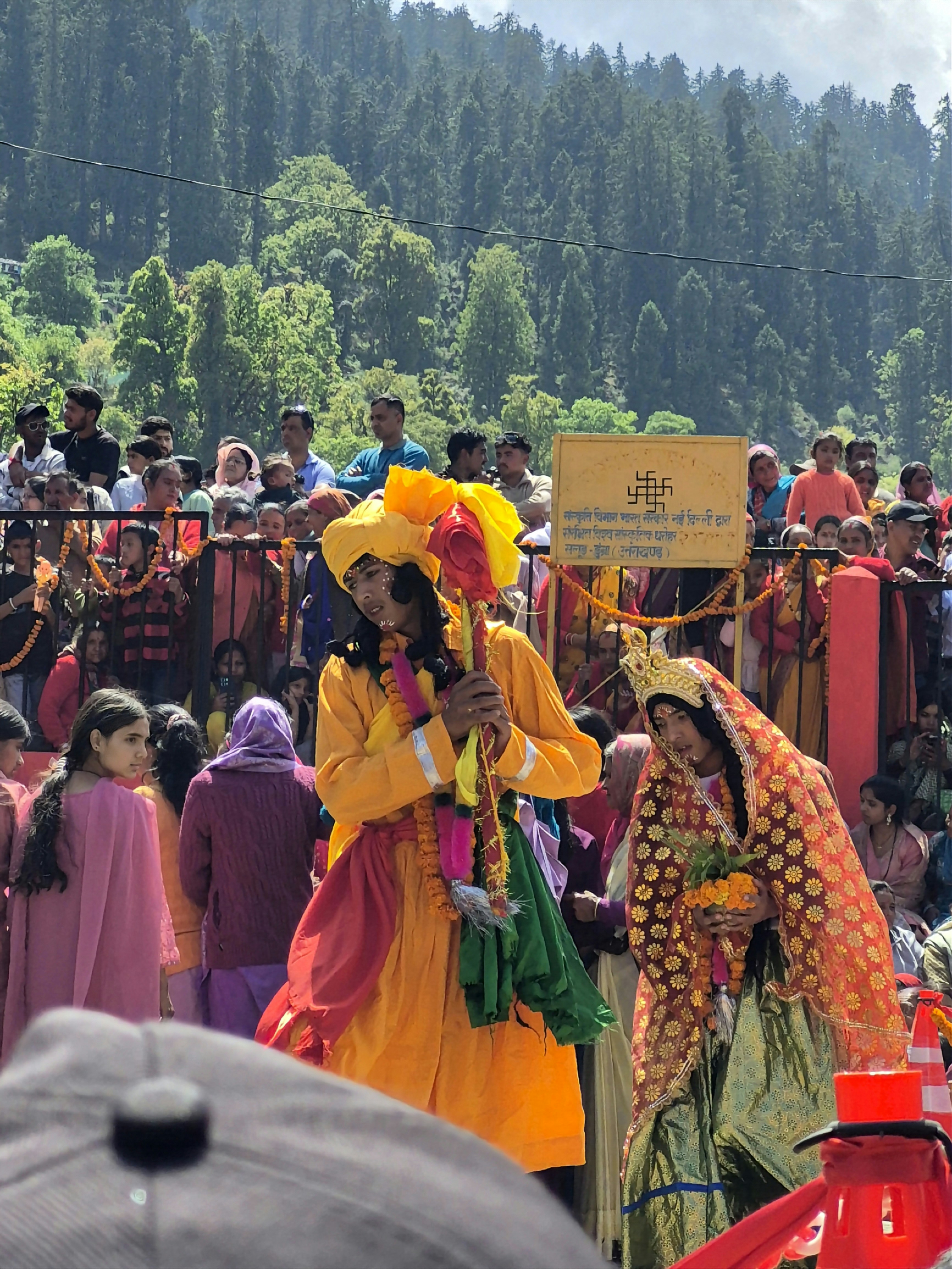 Cultural Performers at UNESCO Ramman Festival in Uttarakhand, India. A stunning close-up shot capturing two local artists in traditional vibrant costumes during the sacred Ramman Utsav in Chamoli. Surrounded by an enthusiastic crowd and set against the lush green pine forests of the Himalayas, this performance brings alive the ancient ritual theater recognized by UNESCO.