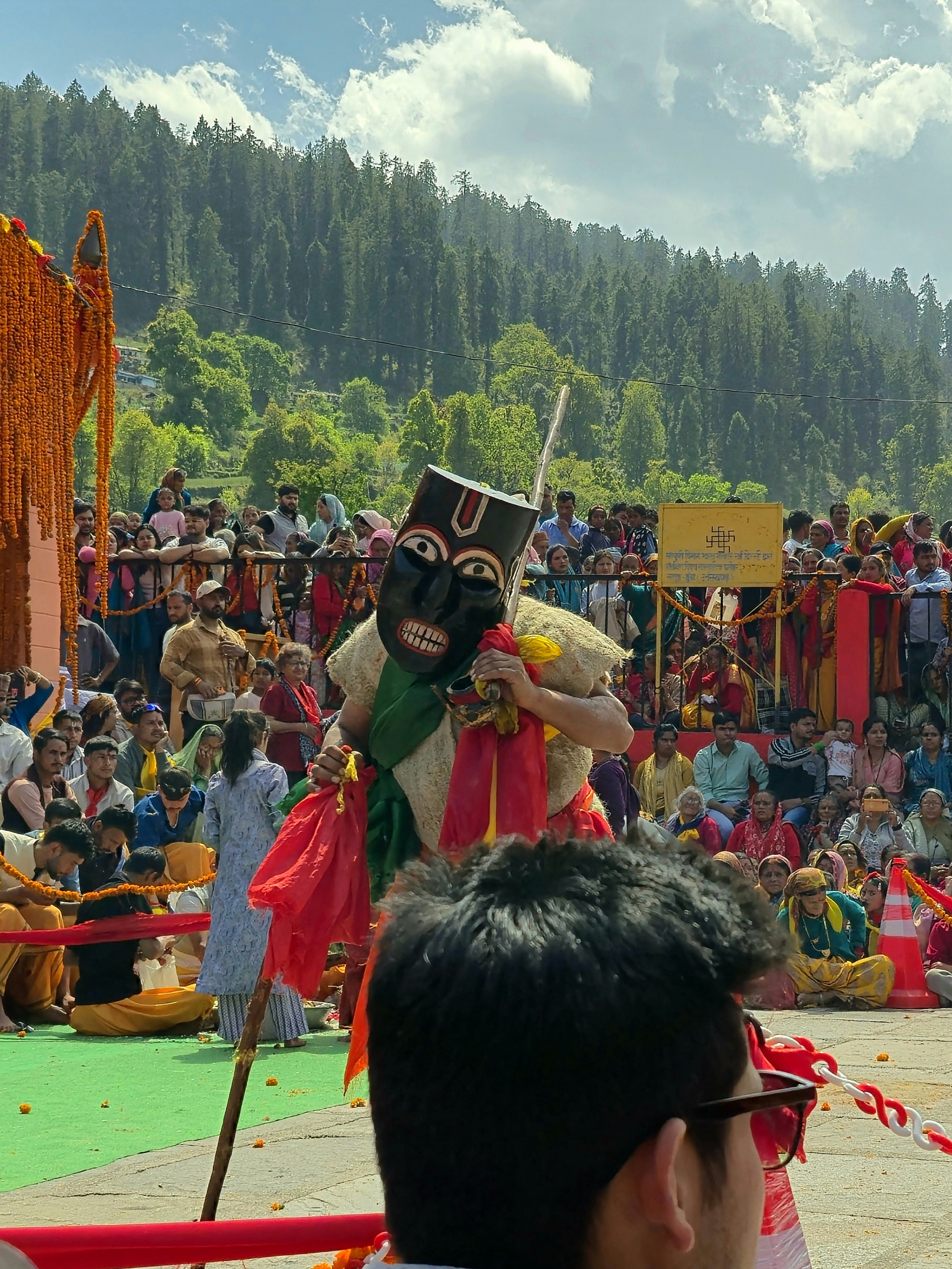 Colorful festival crowd surrounds a central masked performer in a black mask and traditional costume, with a spear in hand, during an outdoor ceremony.