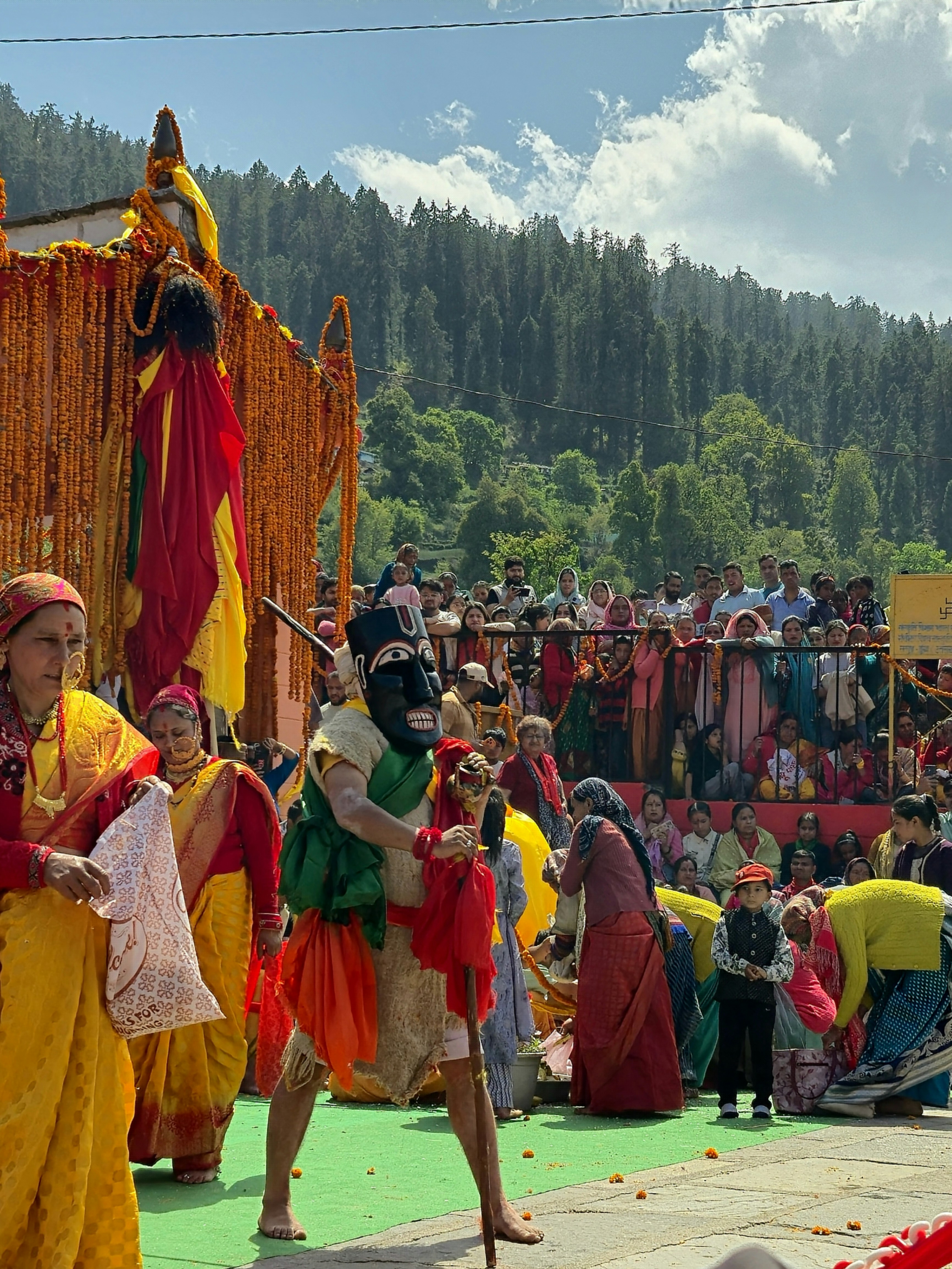 Close-up of Sacred Mask Dance at UNESCO Ramman Festival, Uttarakhand. A brilliant shot capturing a local performer wearing a traditional black ritual mask and holding a staff during the ancient Ramman Utsav in Chamoli, India. The image beautifully blends the colorful presence of local women in traditional yellow attire, an eager crowd of spectators, and the majestic green Himalayan pine forest in the background.