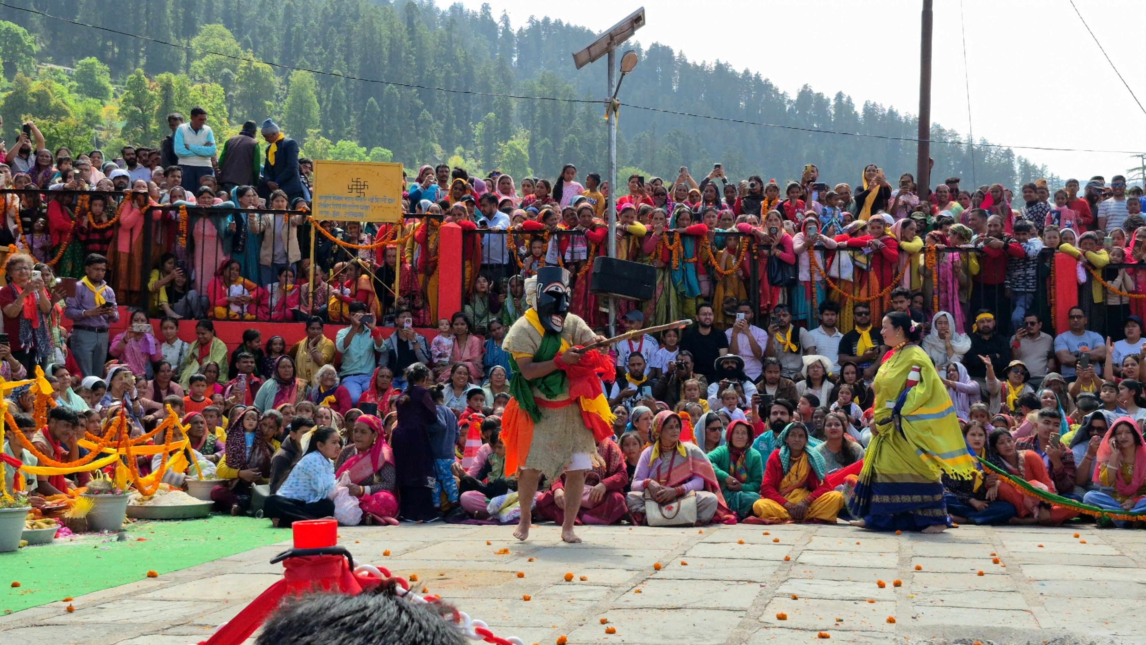 Panoramic View of UNESCO Ramman Mela Festival in Chamoli, Uttarakhand. A spectacular wide landscape shot capturing the grand scale of the ancient Ramman Utsav in India. The image features traditional masked performers enacting a sacred ritual theater on the stone courtyard, completely surrounded by a massive gathering of local villagers, colorful festival decorations, and the majestic Himalayan pine forests under a bright sky.